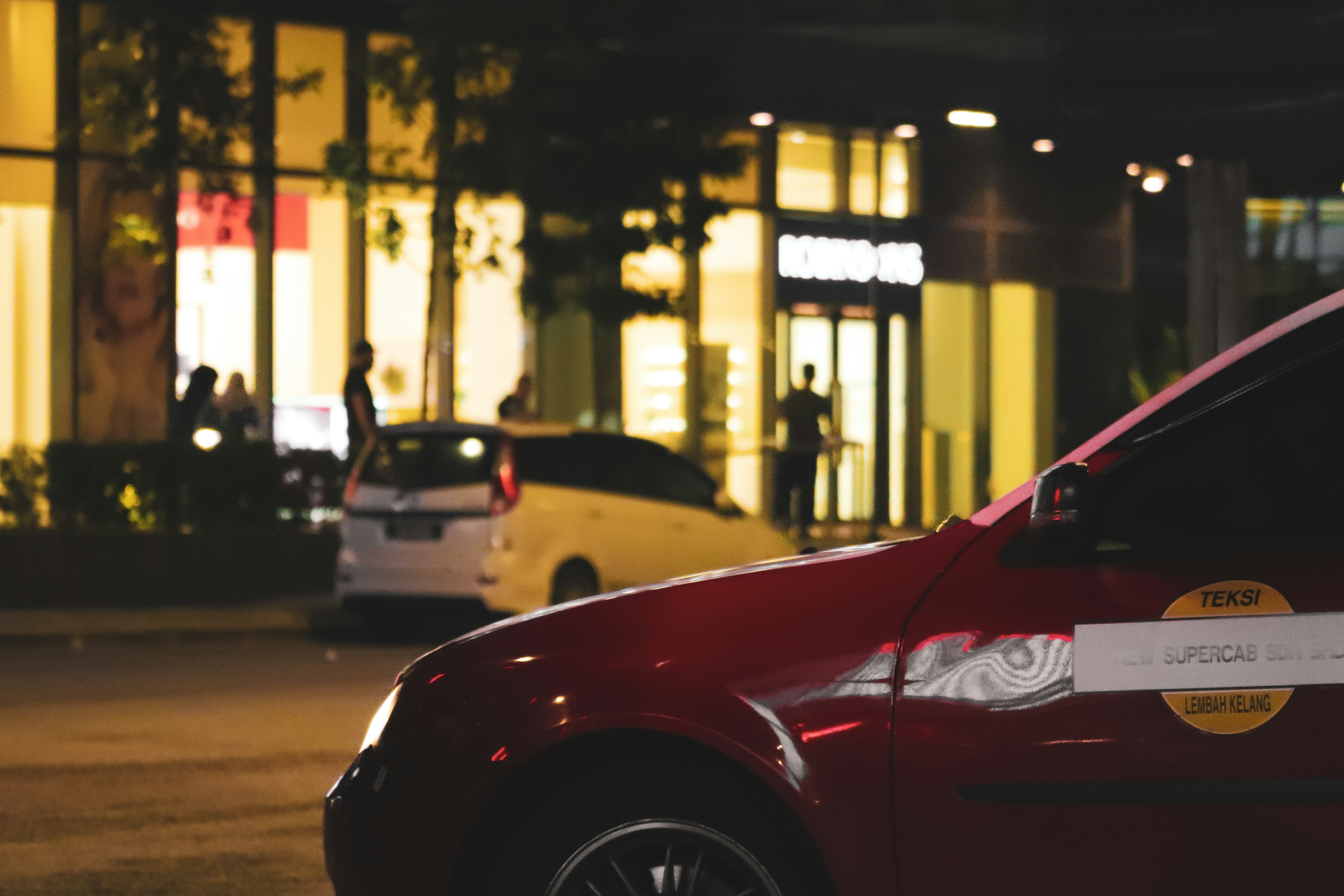 A red taxi parked near a bustling shopping center at night, with illuminated storefronts and silhouettes of people in the background.