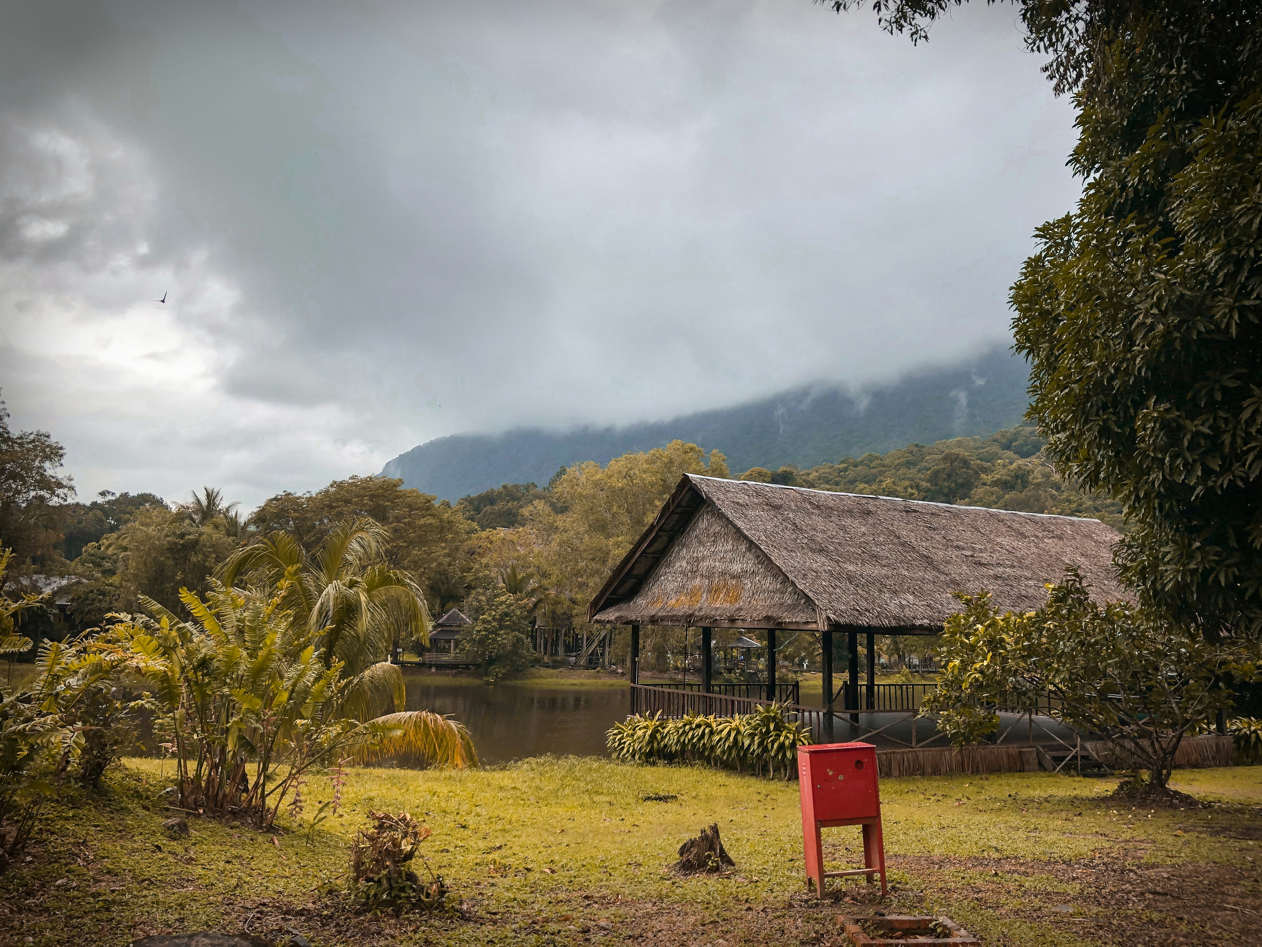Thatched-roof structure beside tranquil pond surrounded by lush greenery and distant mountains under a cloudy sky.