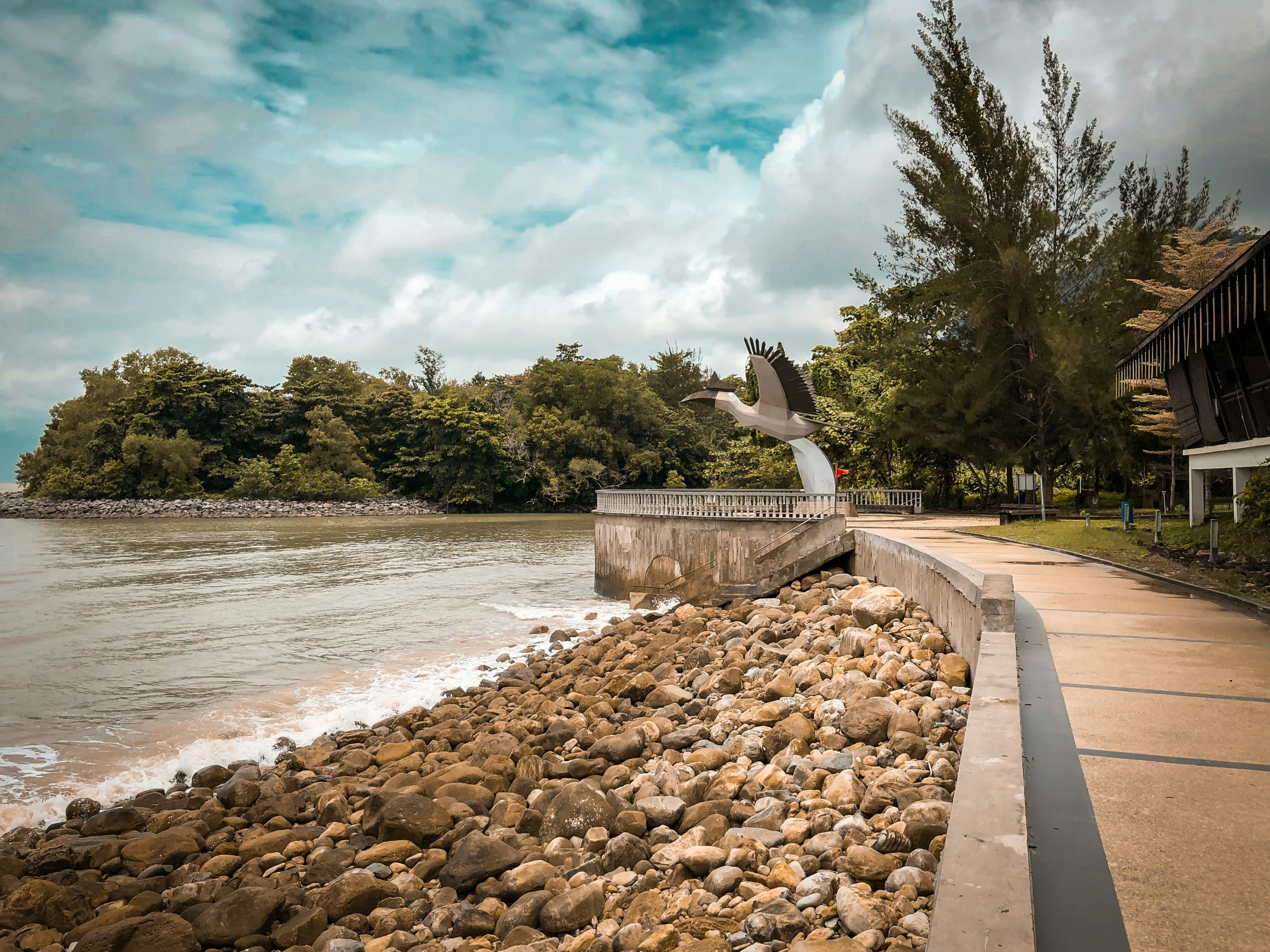 A majestic eagle sculpture stands watch over a rocky shoreline, framed by lush greenery and a tranquil sea under a cloudy sky.