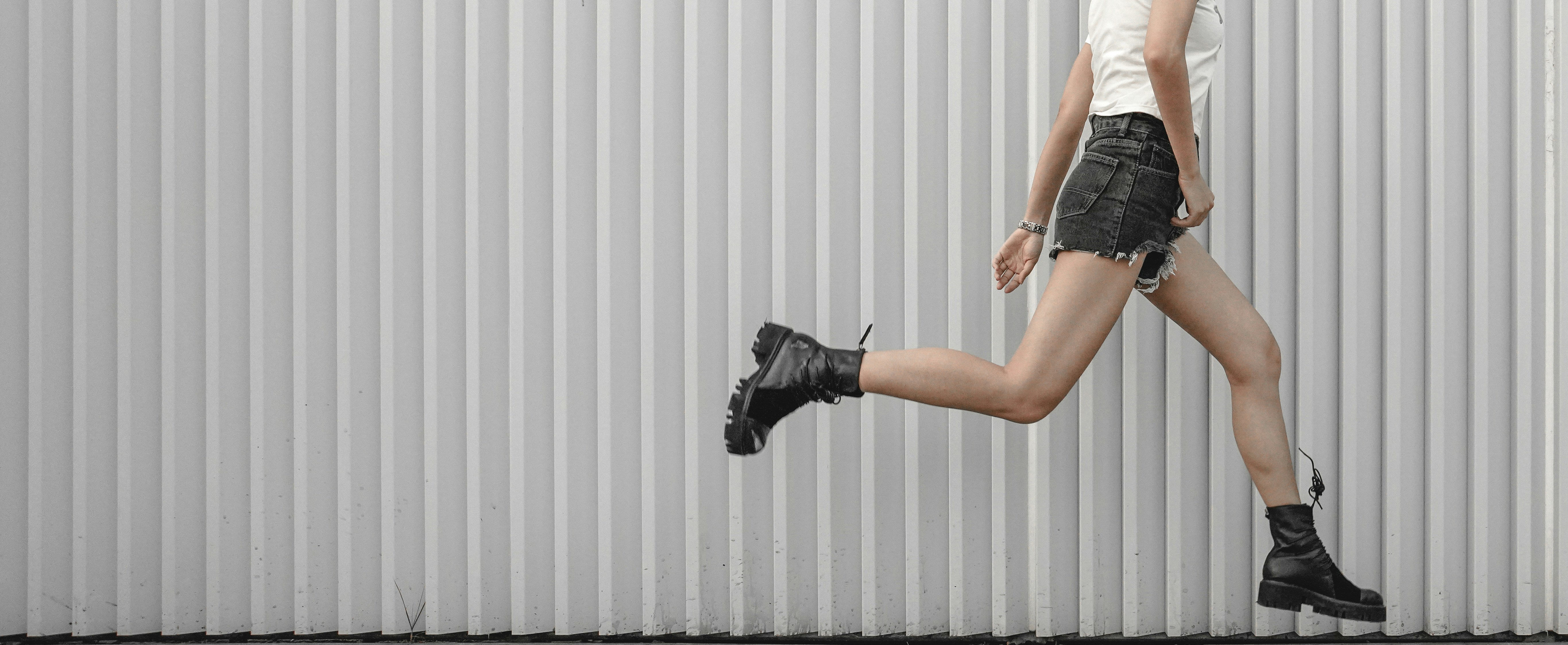 Person in motion wearing black denim shorts and boots against a corrugated metal backdrop.
