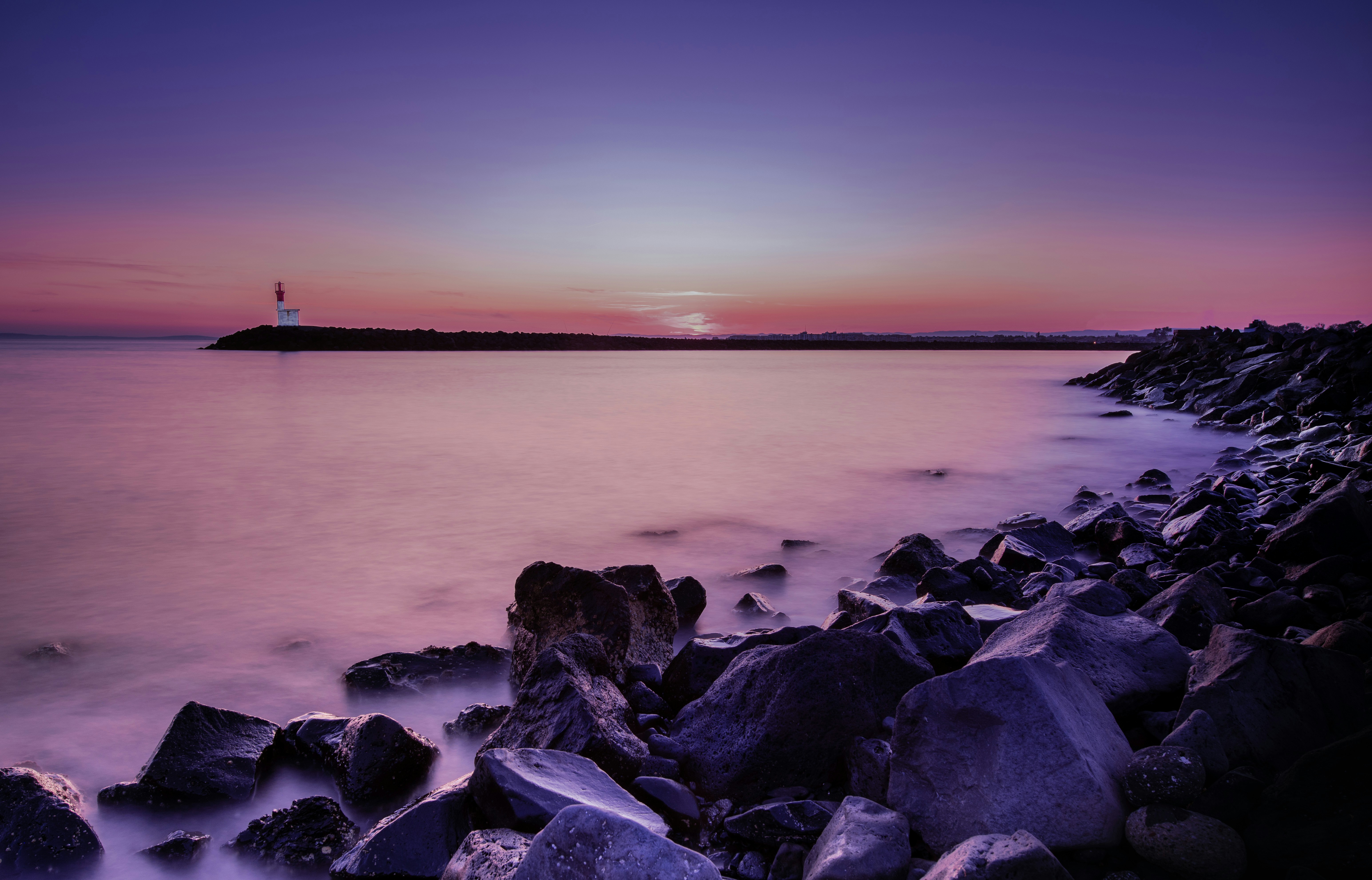 Gentle waves lap against a rocky shore as twilight descends, with a lighthouse standing sentinel in the distance.