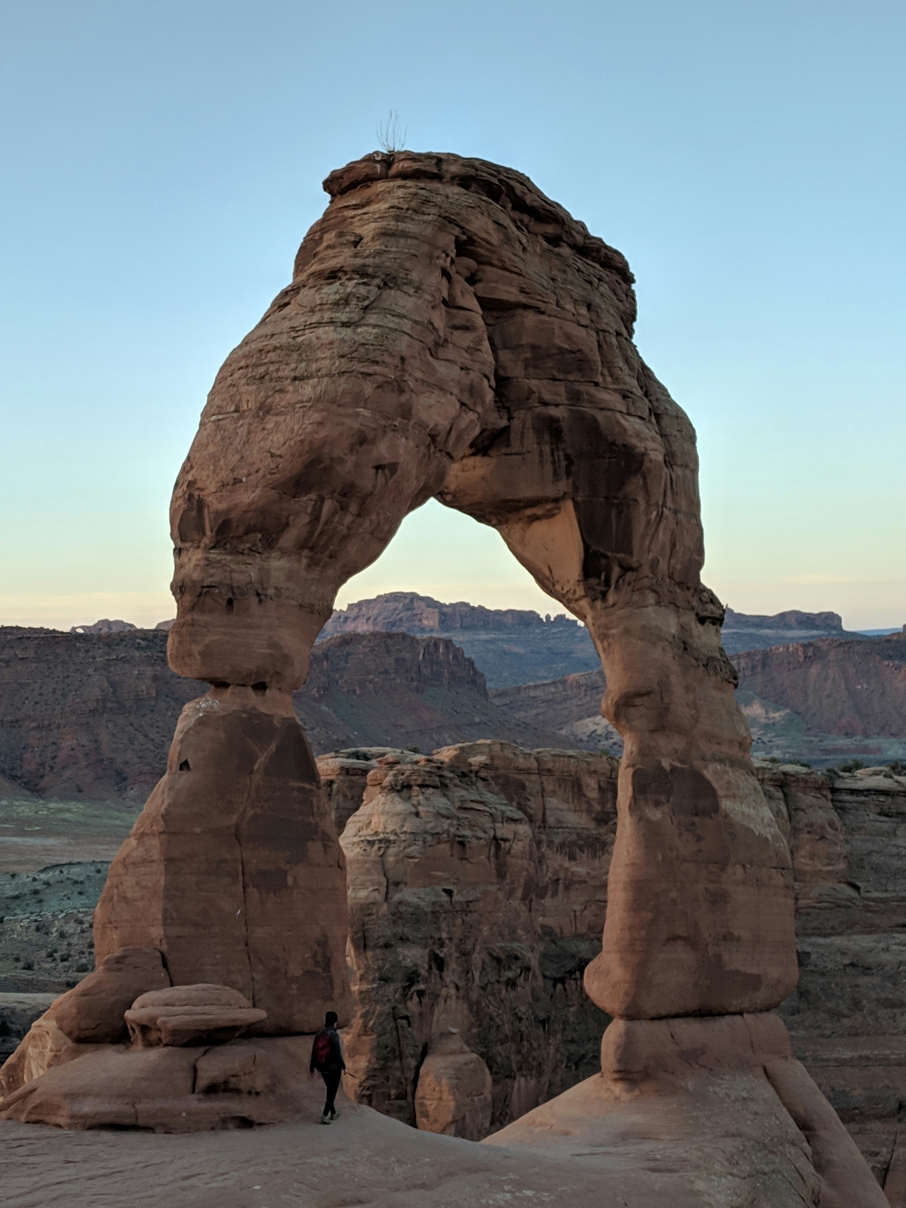 Delicate Arch stands majestically against a backdrop of rugged cliffs and a fading sky, with a lone figure exploring its grandeur.