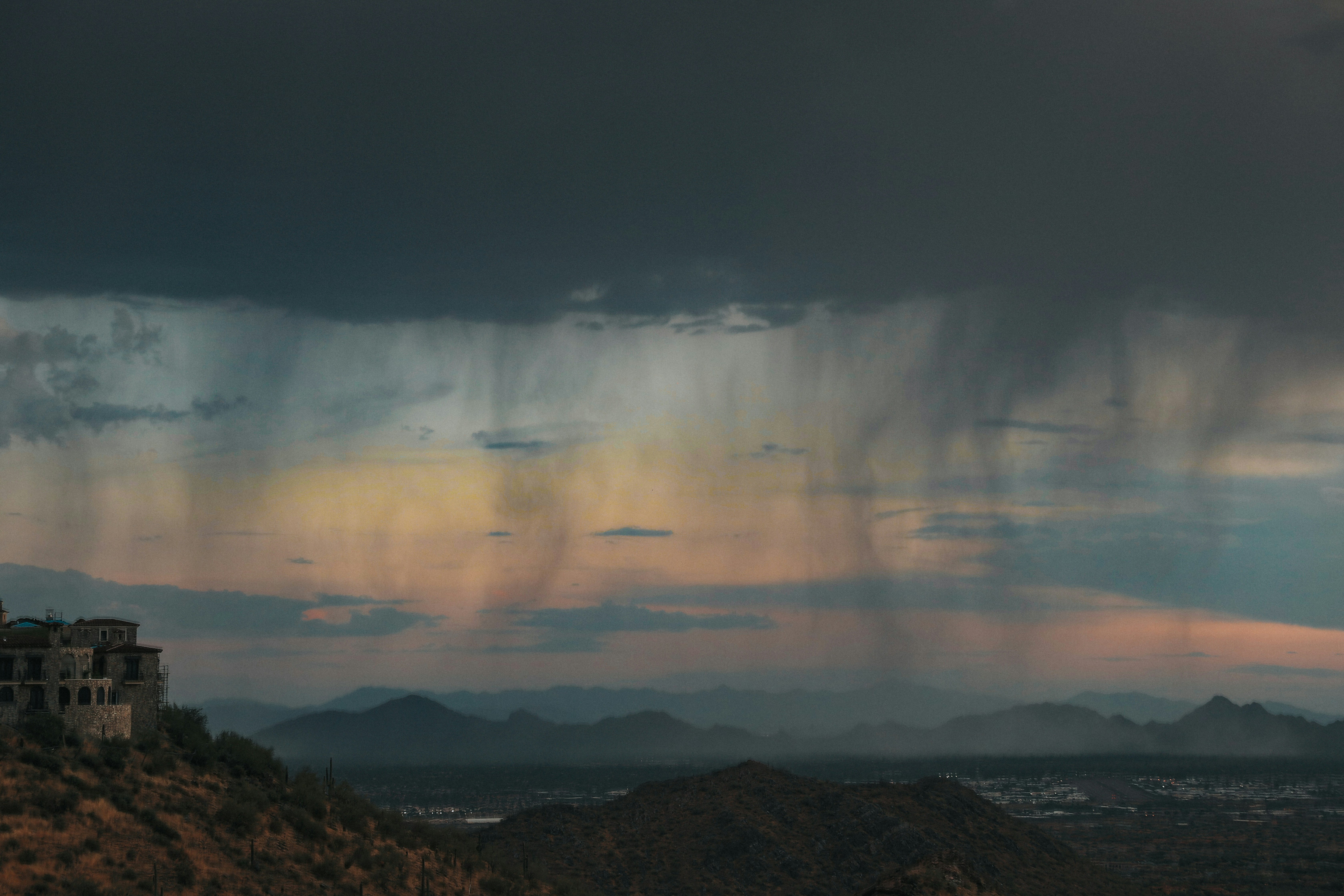 Rainfall streaks descend from dark clouds, illuminating the desert landscape at dusk. A distant structure adds depth to the serene scene.
