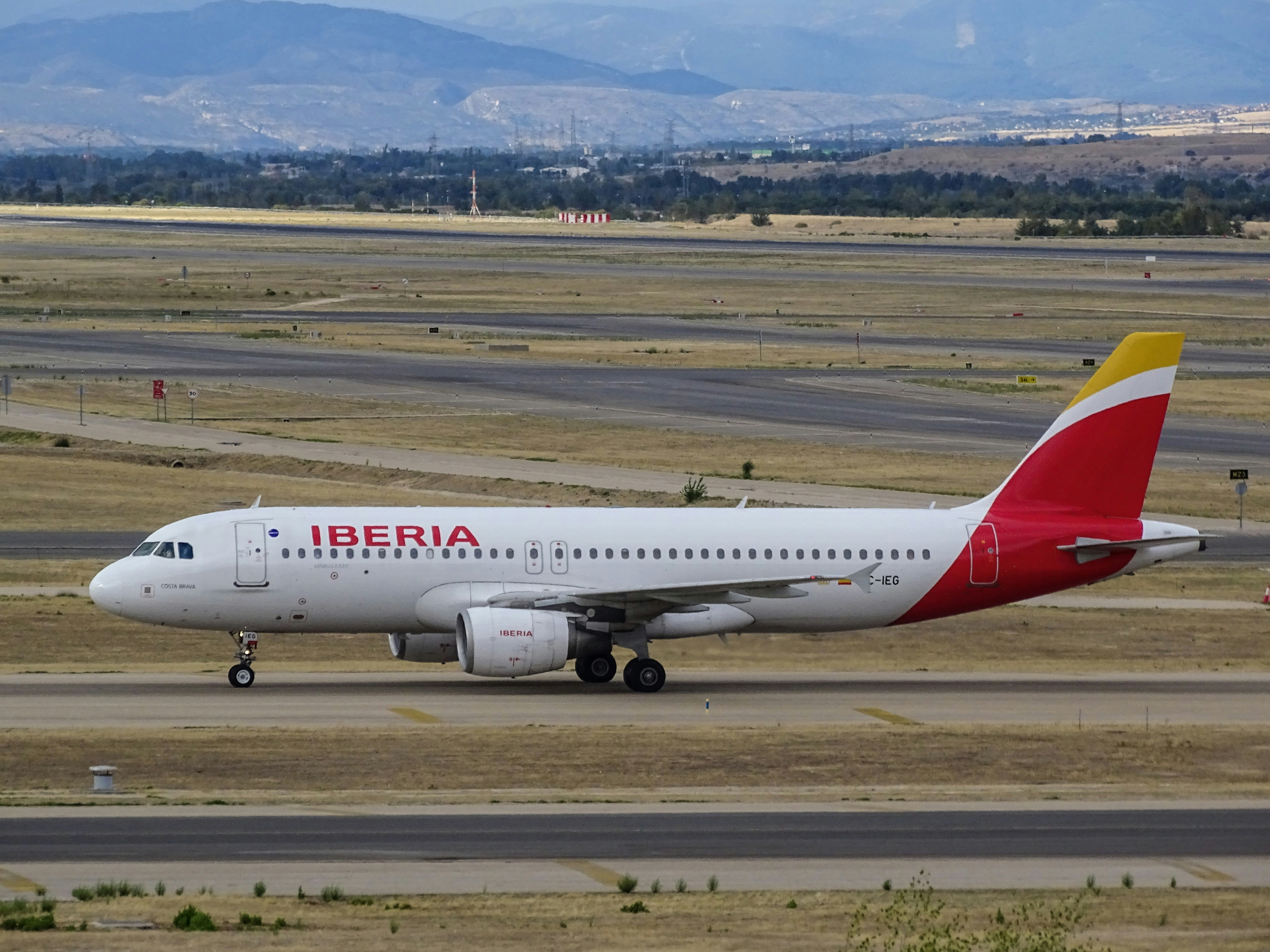 Iberia aircraft taxiing on the runway against a backdrop of distant mountains and clear skies.