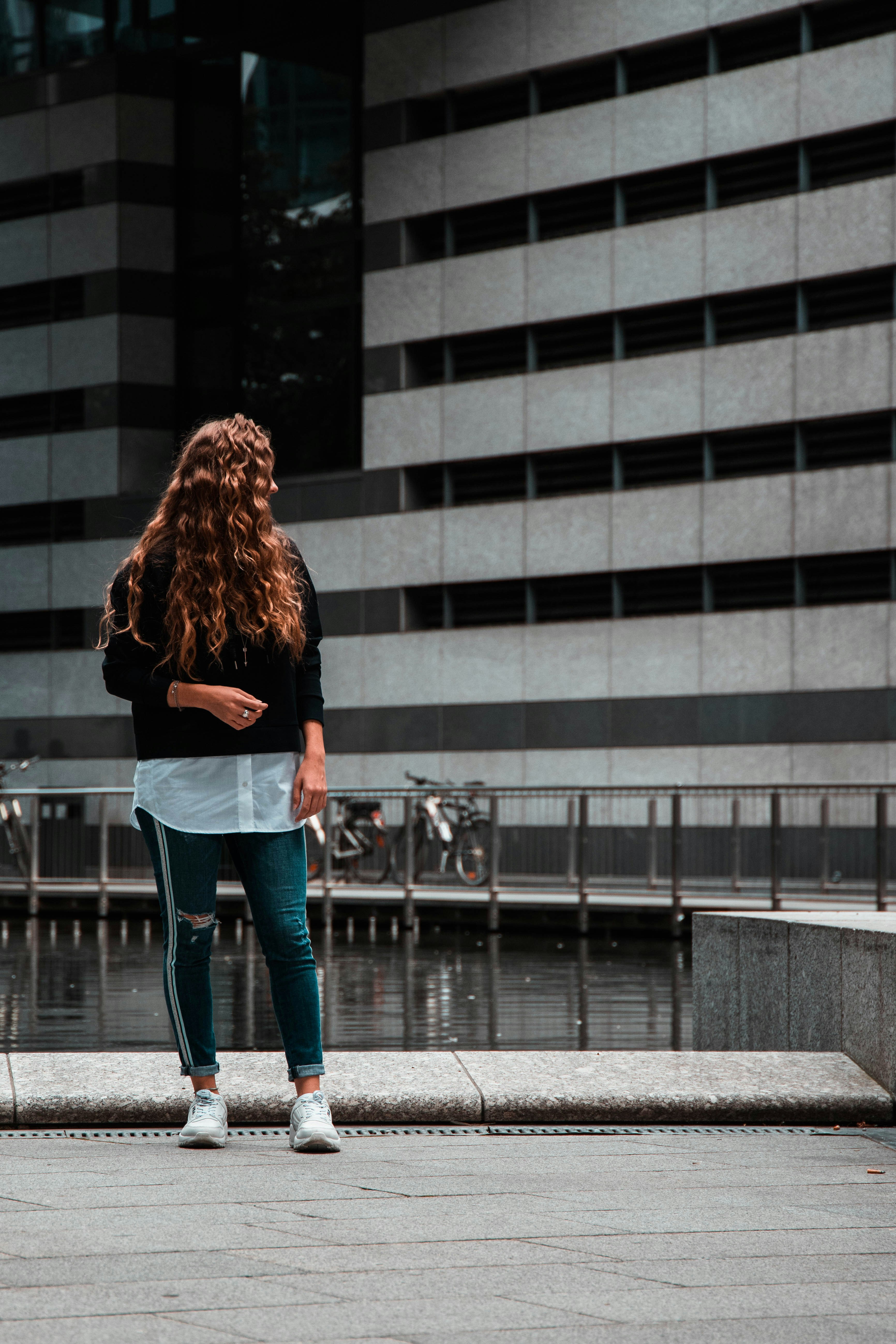 Young woman stands thoughtfully by a reflective pool in an urban setting, framed by modern architecture. Her casual attire contrasts with the sleek surroundings.