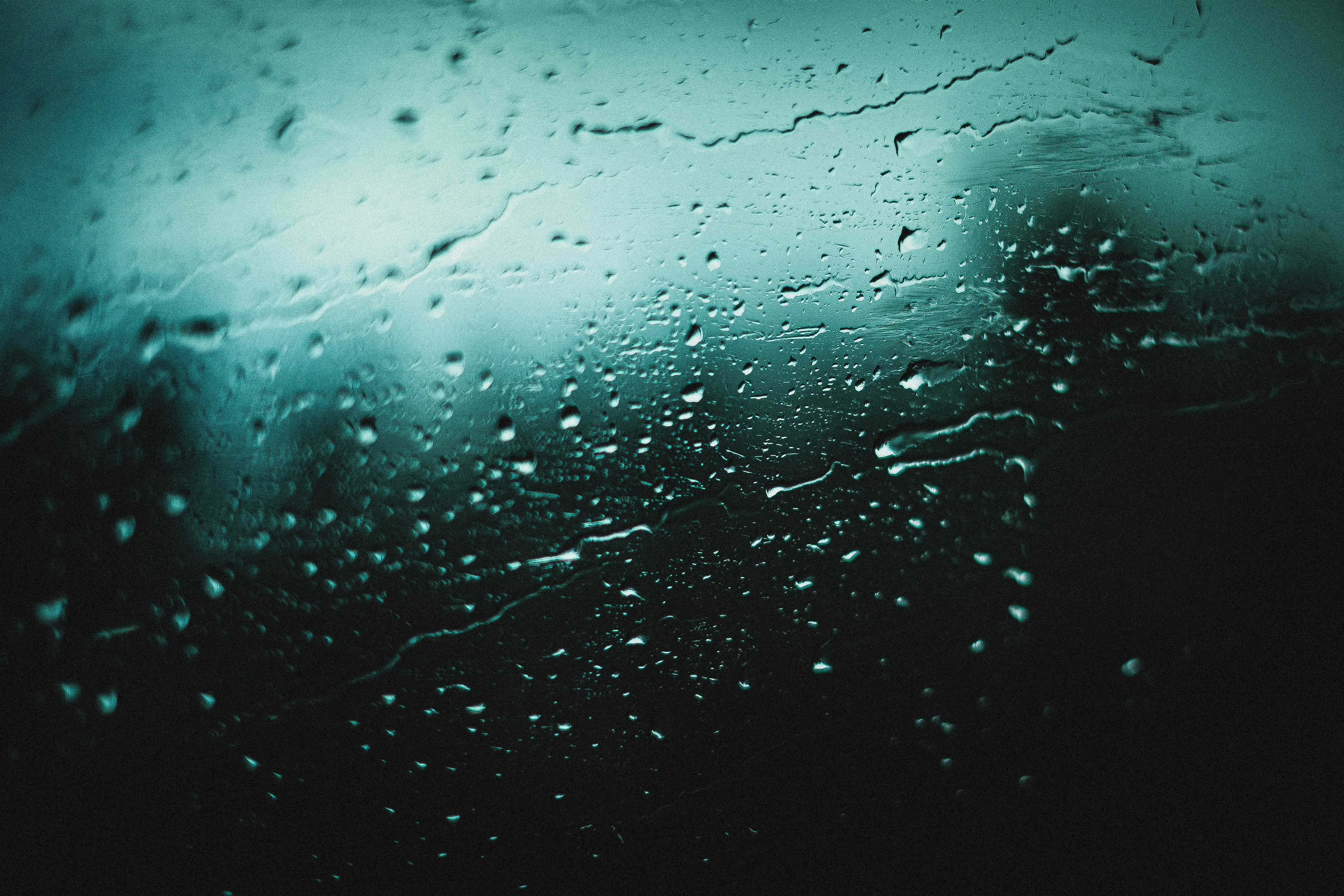 Close-up of raindrops cascading down a misty window, creating a moody atmosphere with blurred silhouettes in the background.