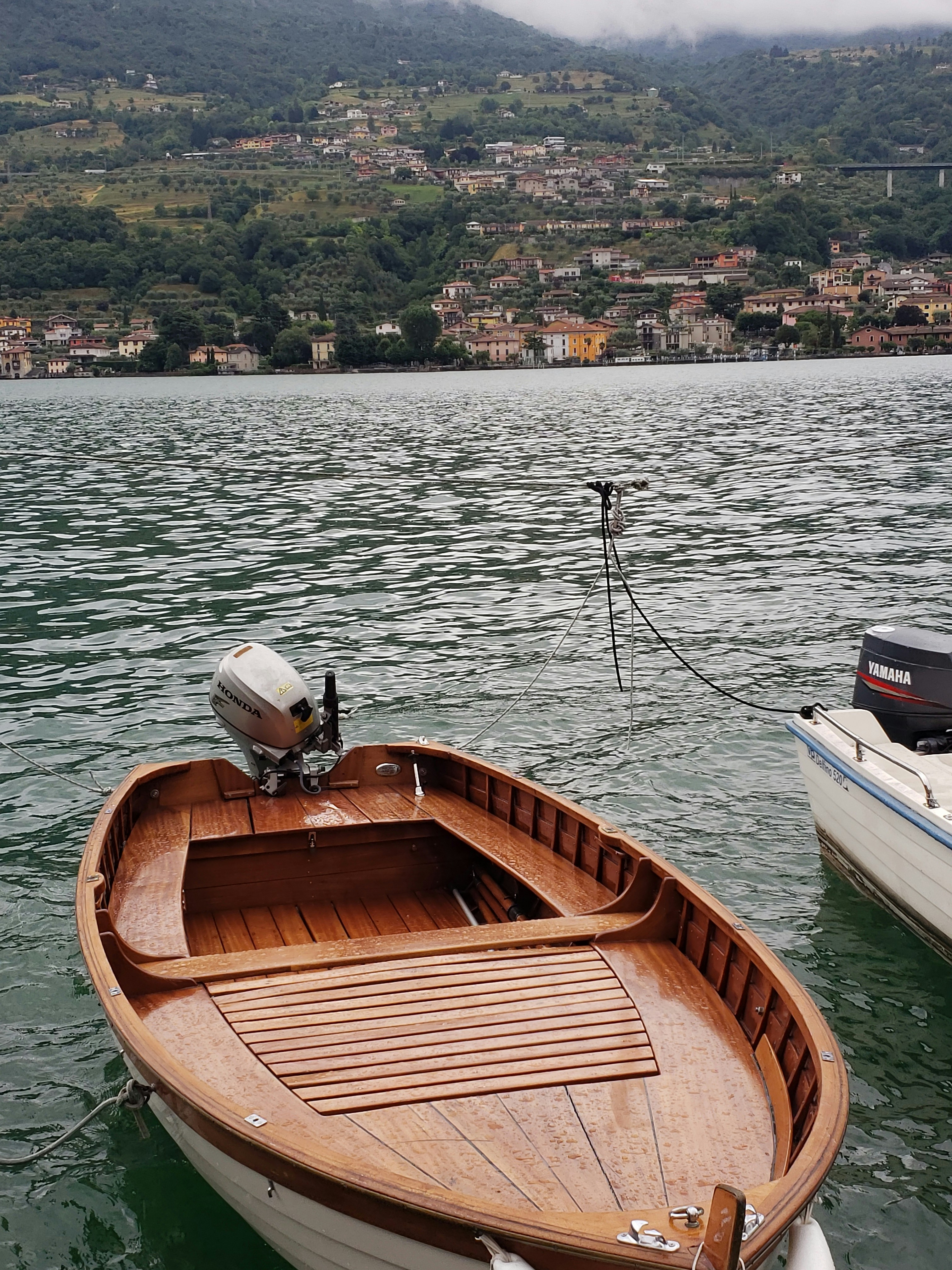 A wooden boat anchored in tranquil waters, showcasing its intricate craftsmanship against a picturesque lakeside backdrop. The gentle ripples reflect the surrounding landscape.