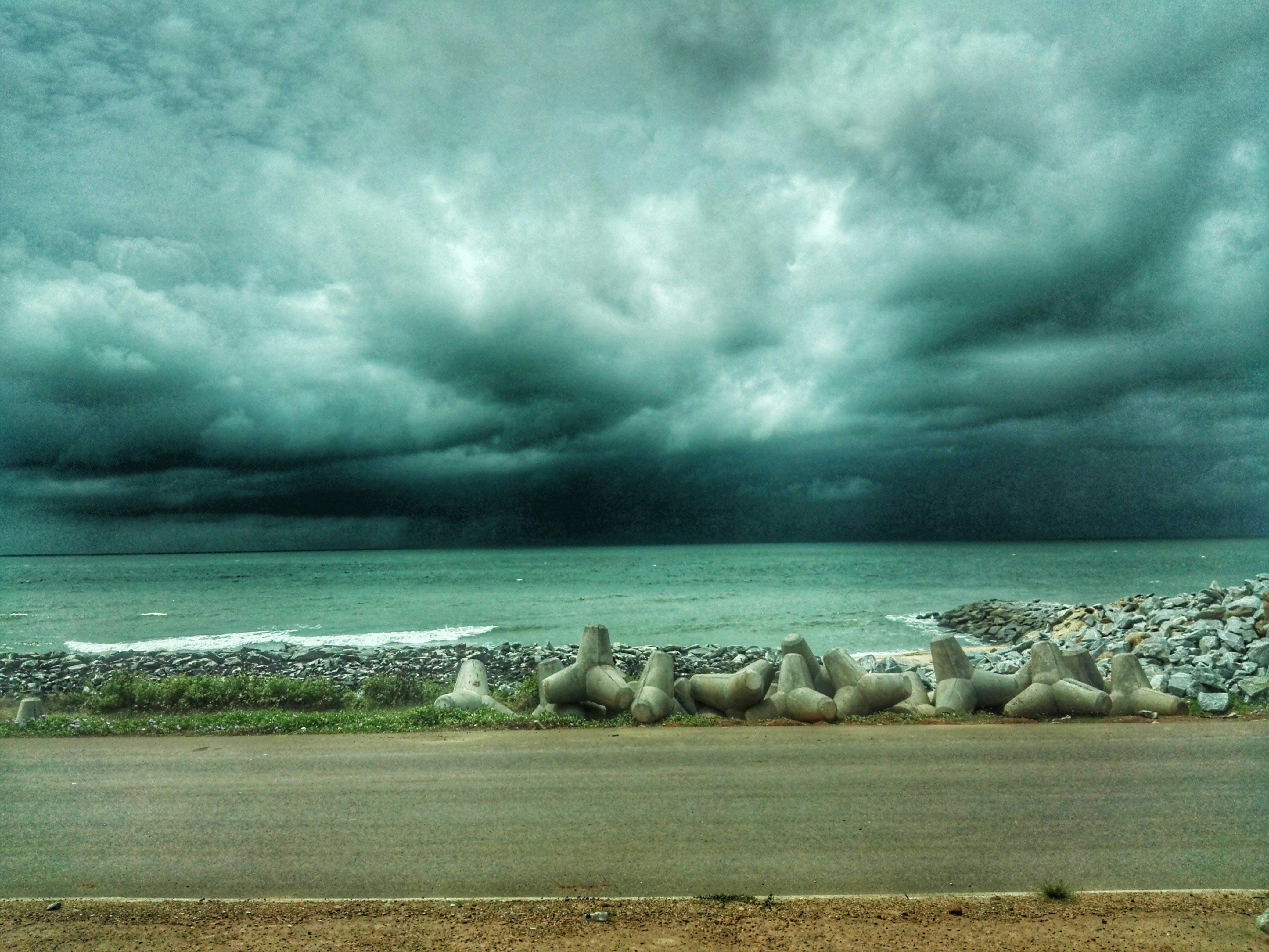 Storm-laden turquoise sea meets a tetrapod breakwater along a rocky shoreline beneath an emerald, cloud-laden sky.