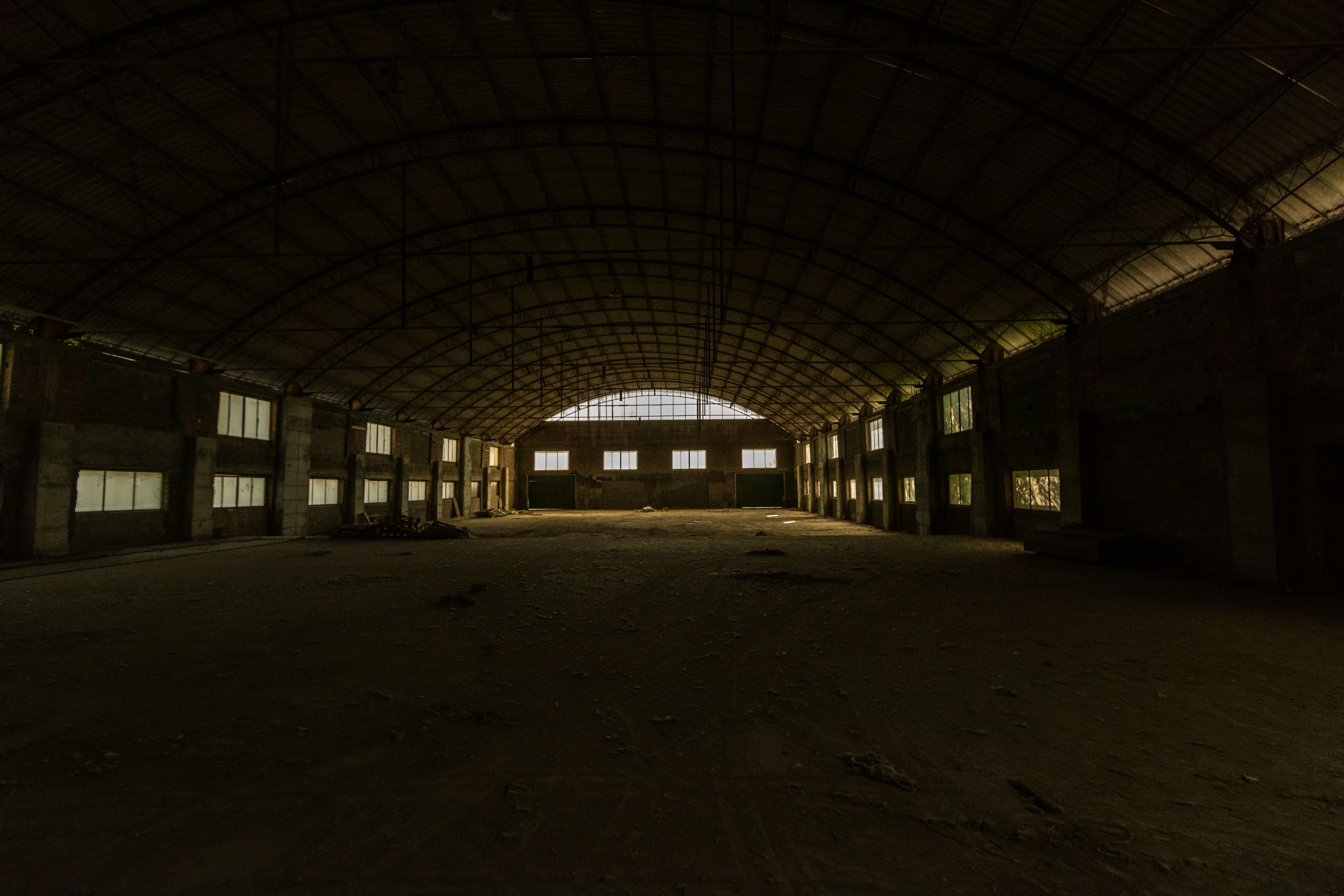 Expansive interior of an abandoned warehouse, featuring high ceilings and dim lighting with scattered debris on the floor.