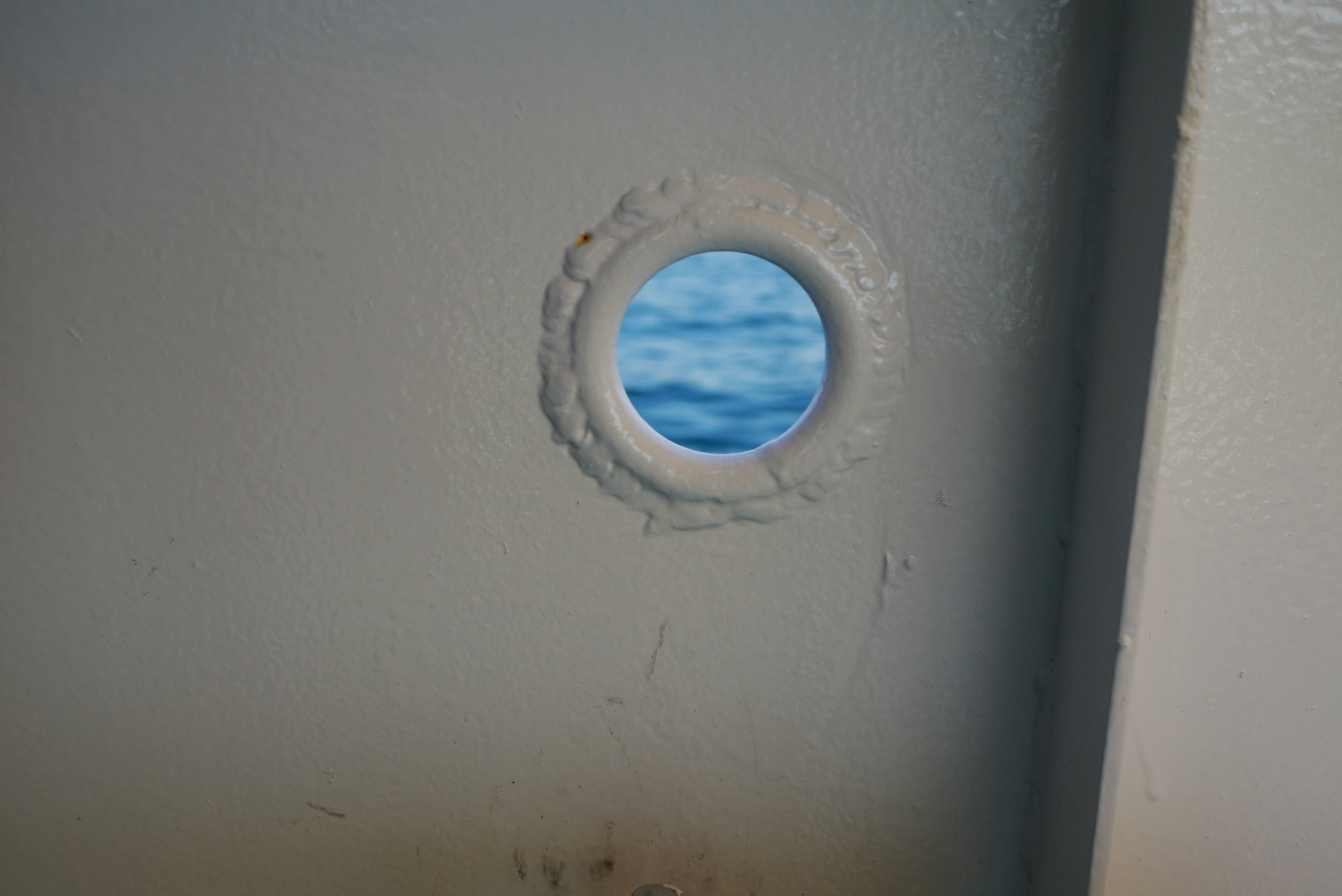 Circular porthole in a painted hull reveals blue water beyond, creating a quiet focal contrast.