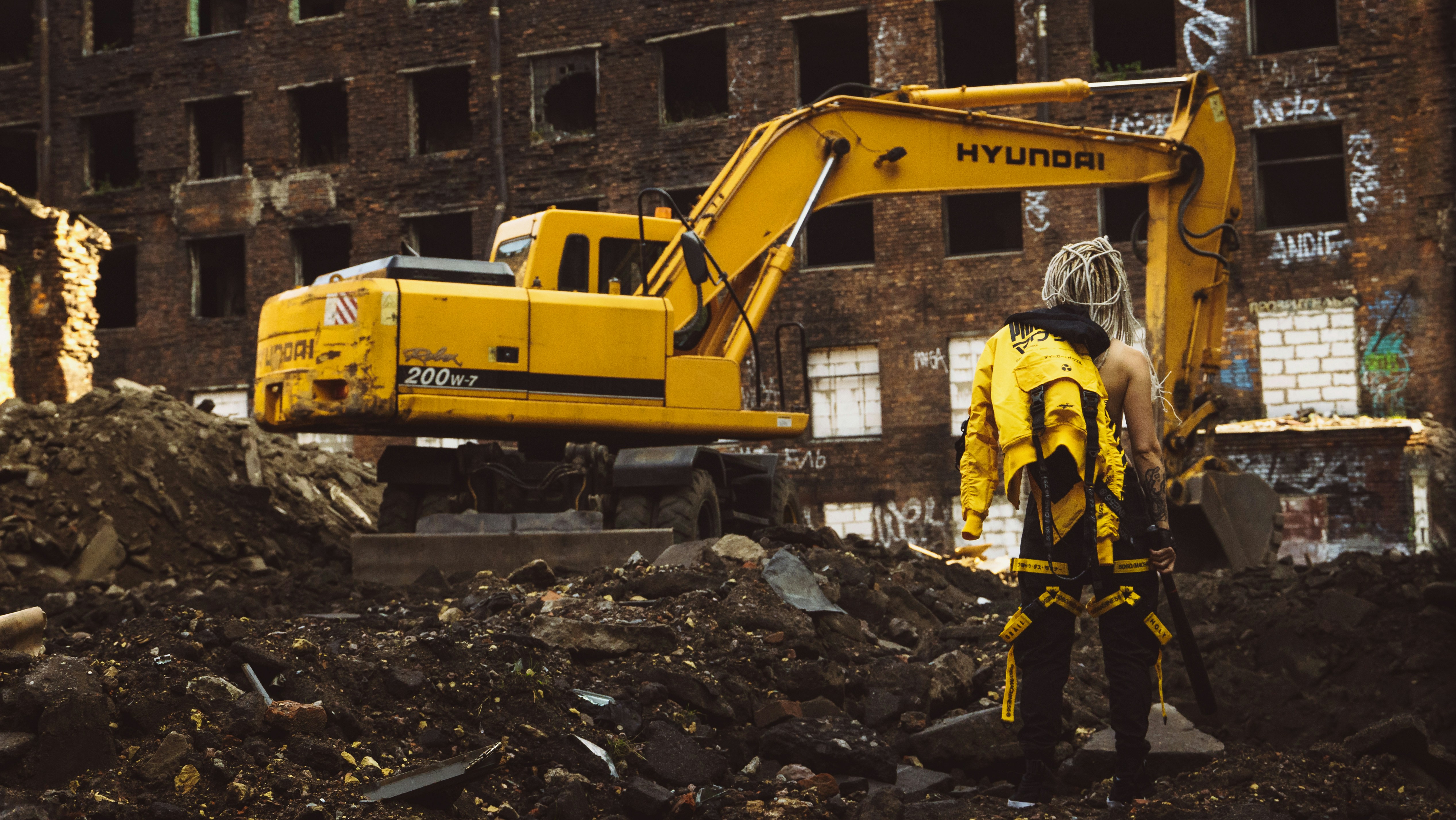A figure in a yellow outfit stands amidst debris, observing a yellow excavator at a derelict construction site. The contrast between human and machinery highlights themes of urban decay and renewal.