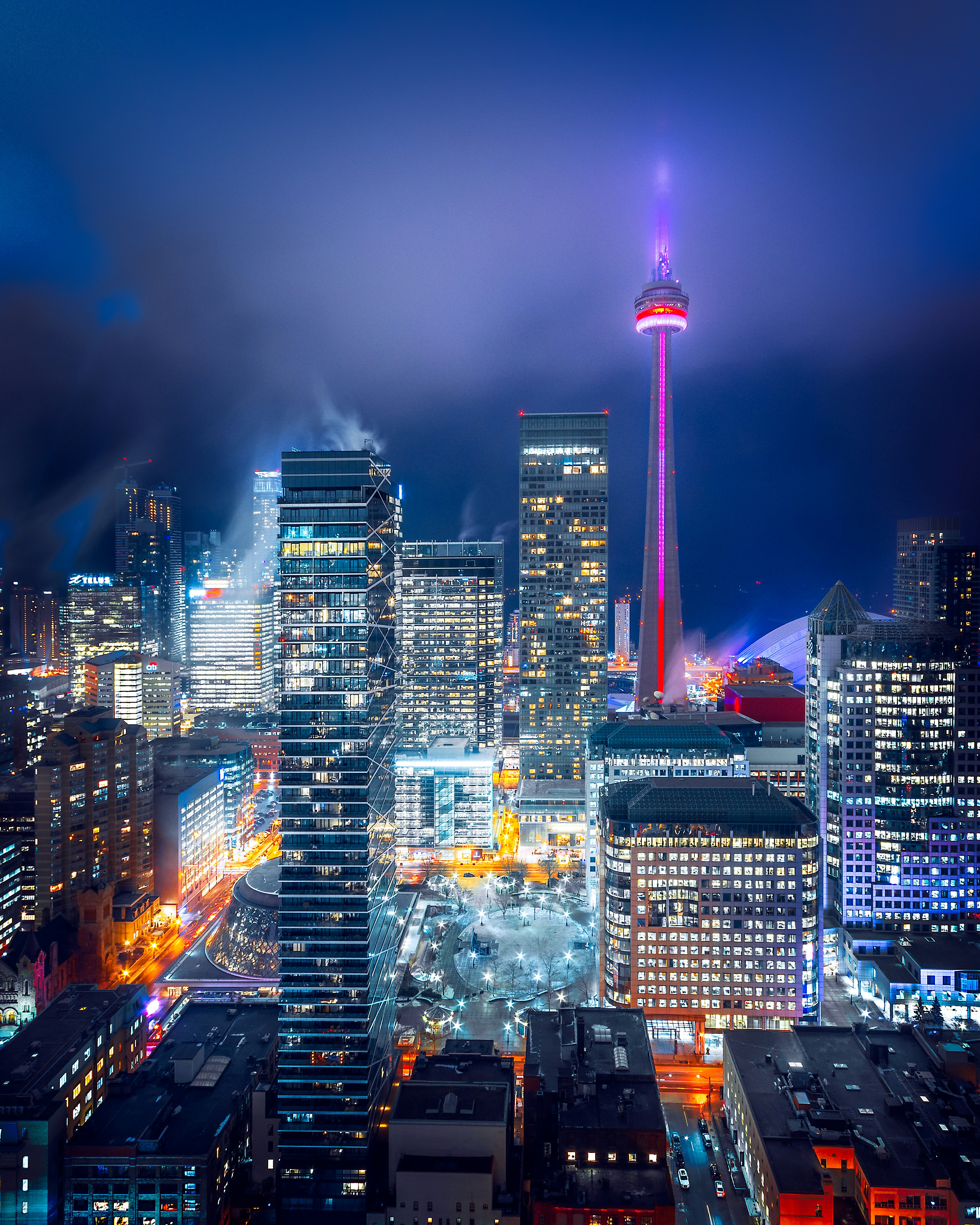 Illuminated skyline of a bustling city at night, featuring the CN Tower prominently lit in vibrant colors. The scene captures the dynamic energy of urban life.