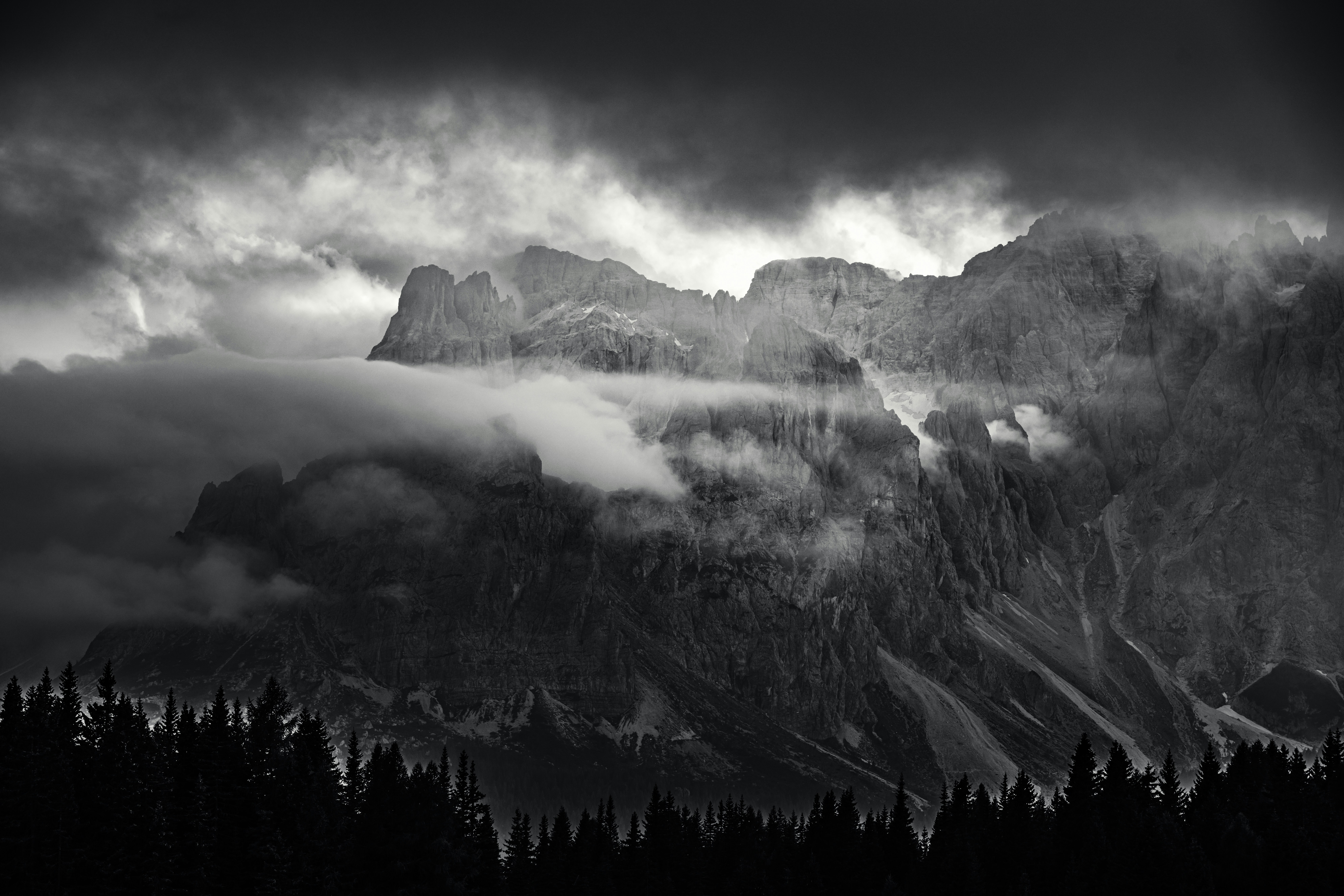 Dramatic clouds envelop rugged mountain peaks in grayscale, with a dense forest in the foreground.