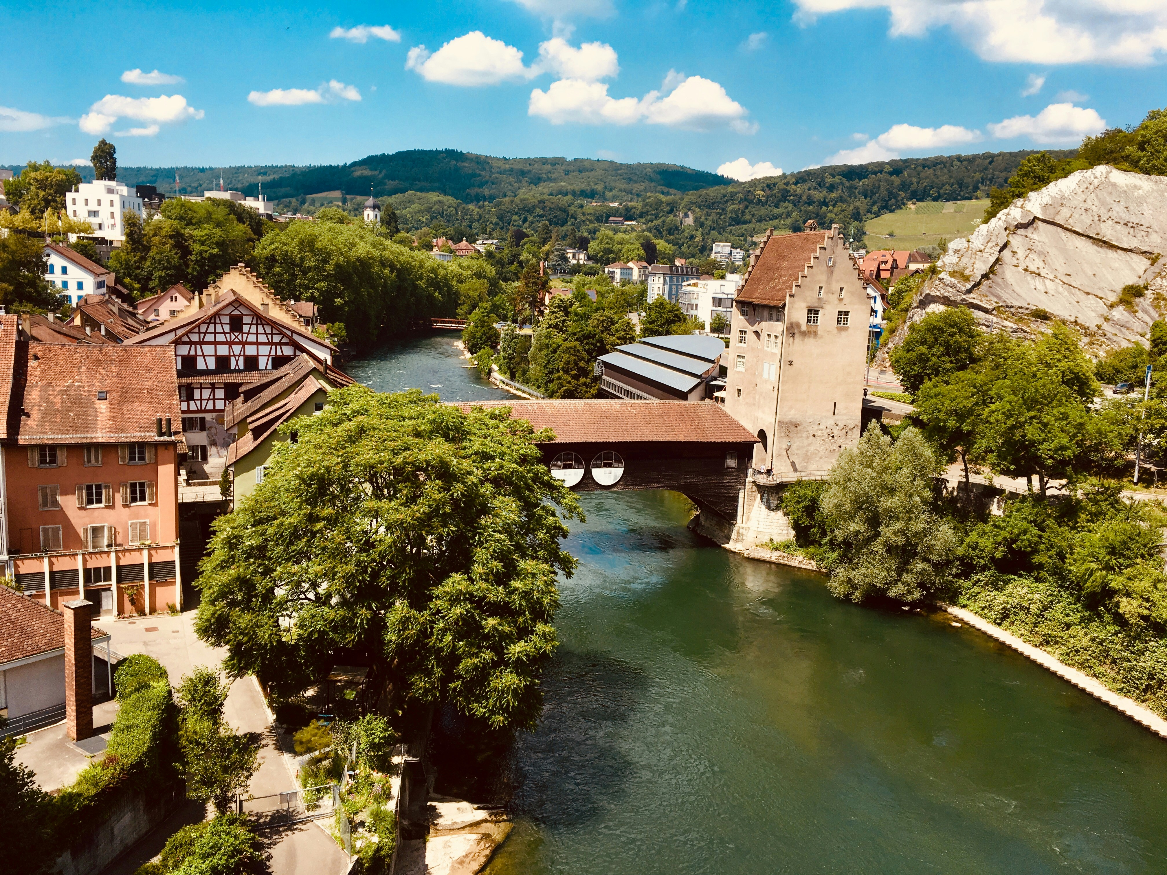 Historic bridge connecting quaint buildings along a river, framed by lush greenery and distant hills.