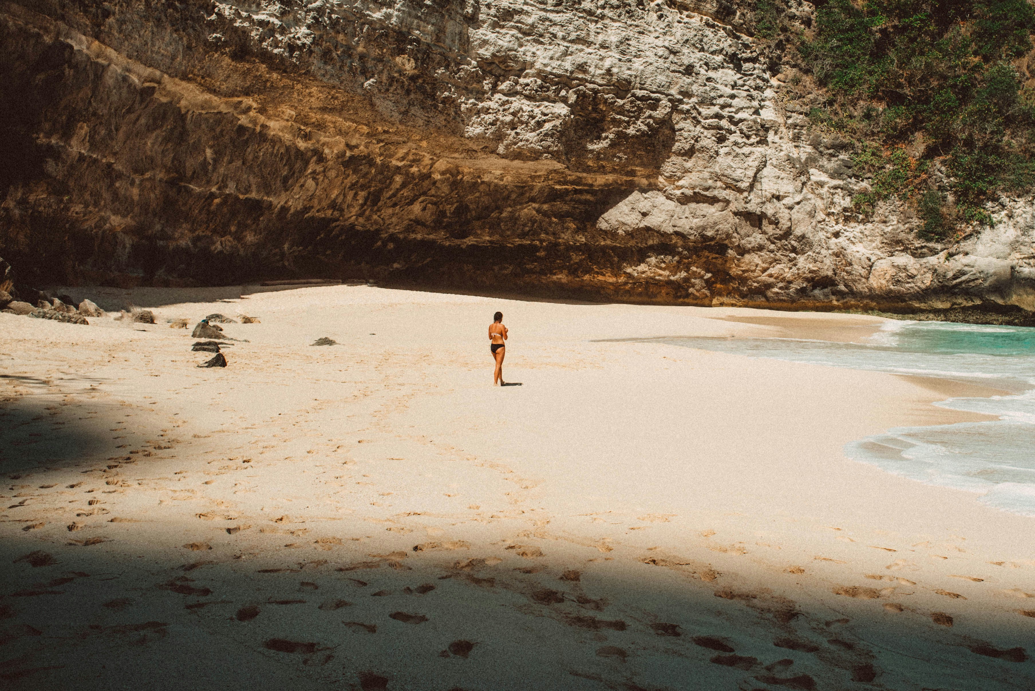 Person standing on a quiet beach with towering cliffs and gentle waves under soft sunlight.
