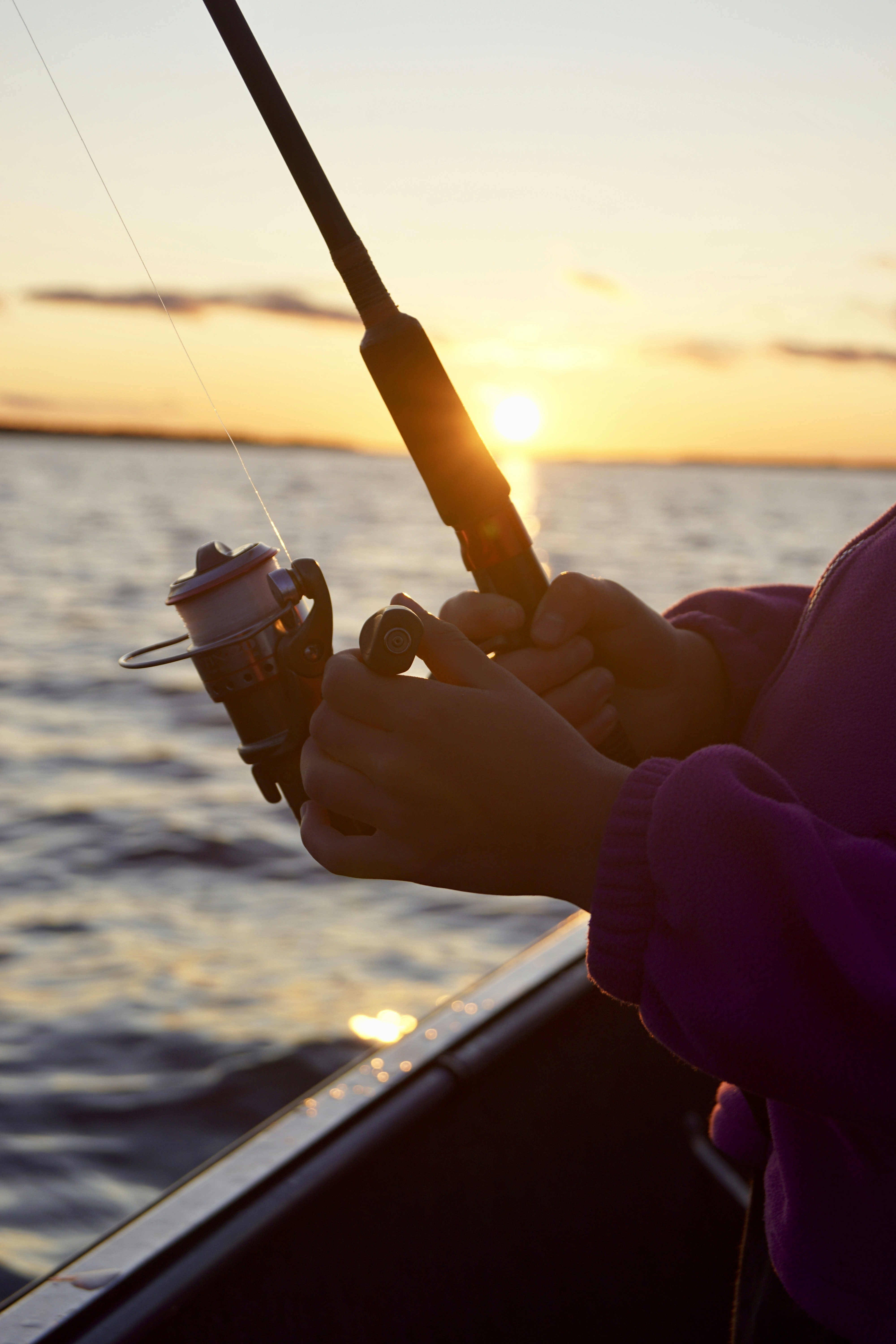 Hands gripping a fishing rod, silhouetted against a vibrant sunset over water. The scene captures the tranquility of fishing as day transitions to night.