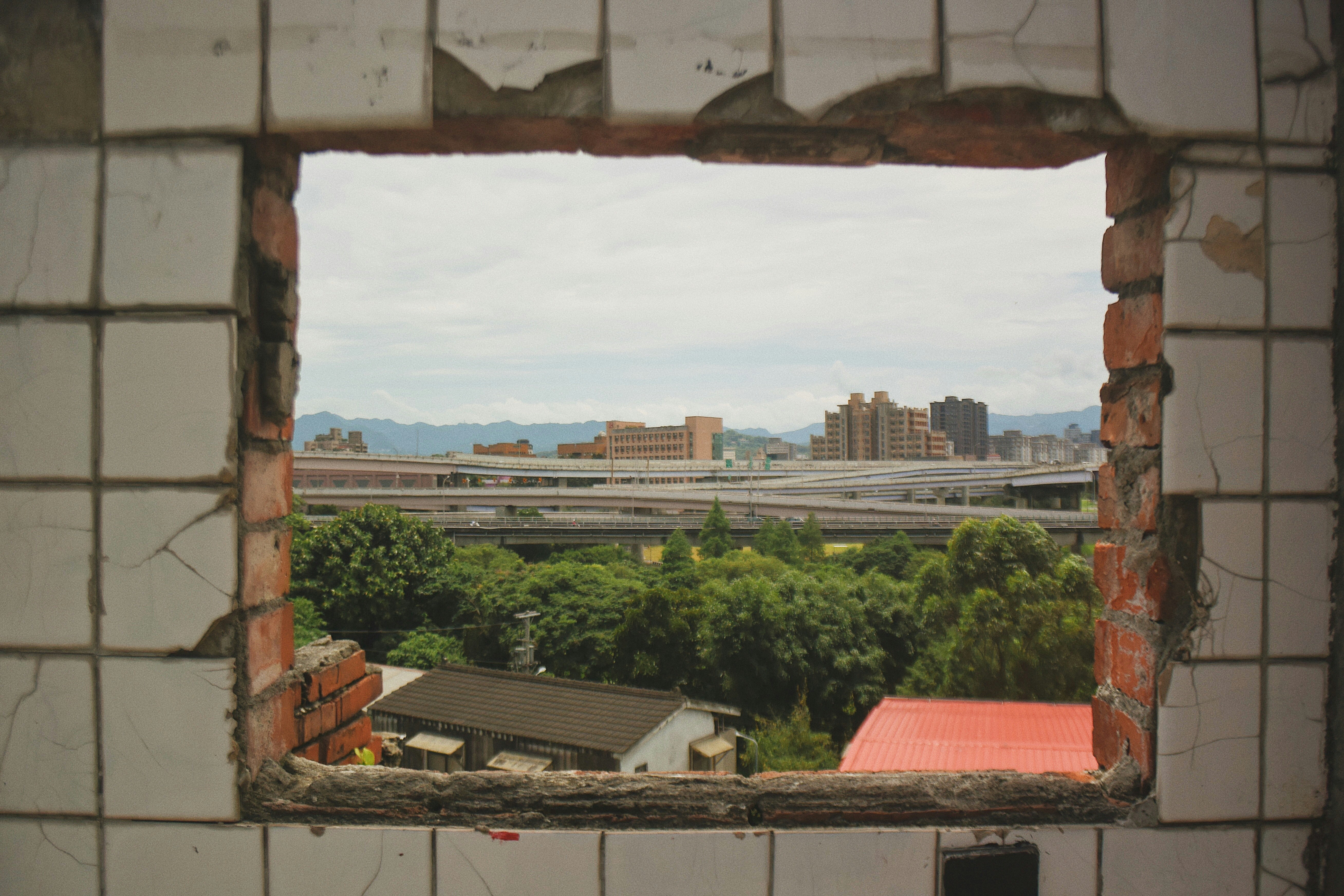 Cityscape seen through a crumbling tile-framed window, with trees in the foreground and a distant skyline.