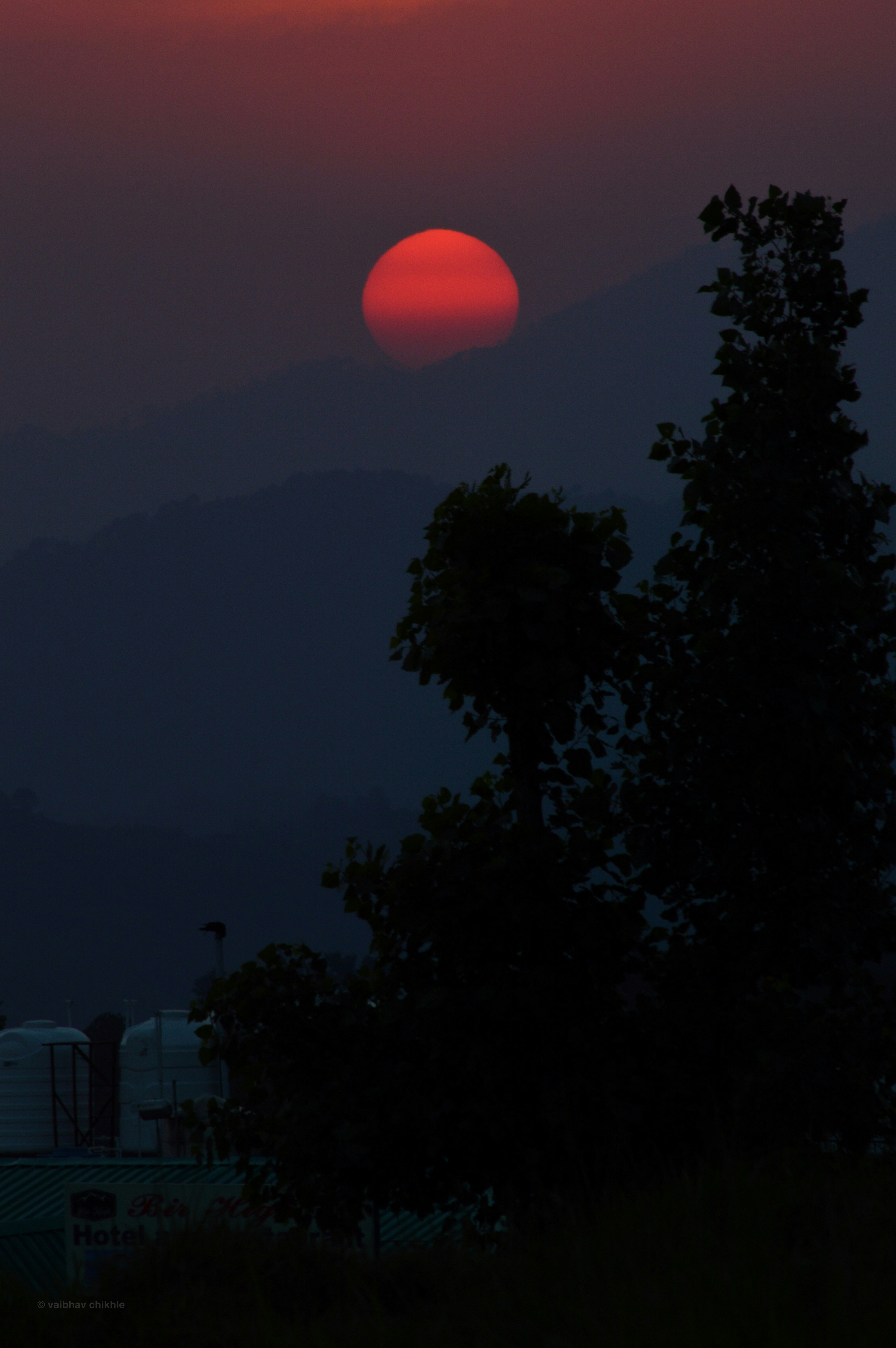 Vibrant red sun setting behind dark mountain silhouettes, casting a serene atmosphere. The foreground features shadowy trees adding depth to the scene.