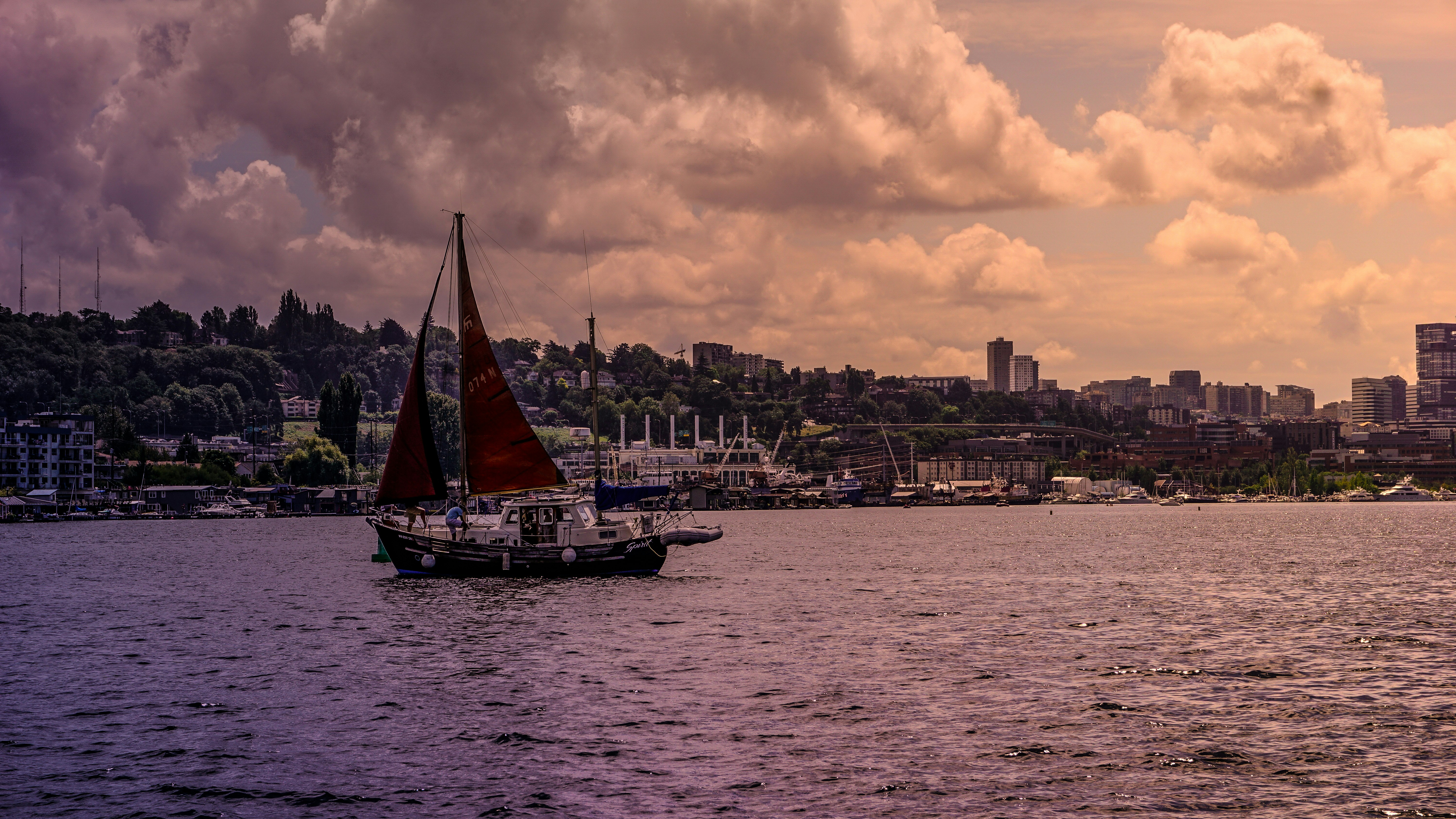 Sailboat with red sails gliding across a harbor under a dramatic, cloud-filled sky at sunset.