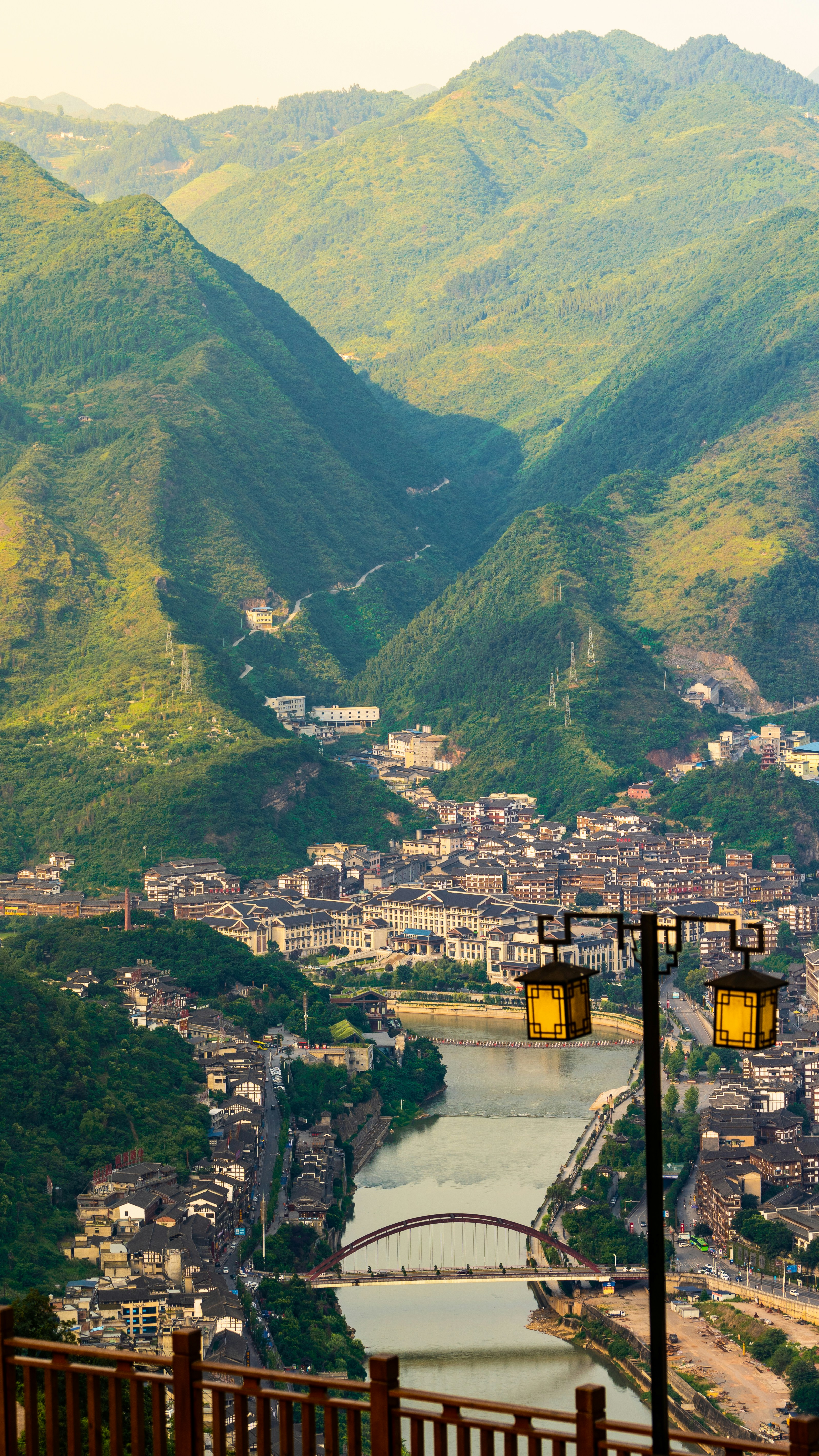 A panoramic view of a serene river winding through a quaint town nestled in lush mountains, accentuated by traditional architecture and modern infrastructure.