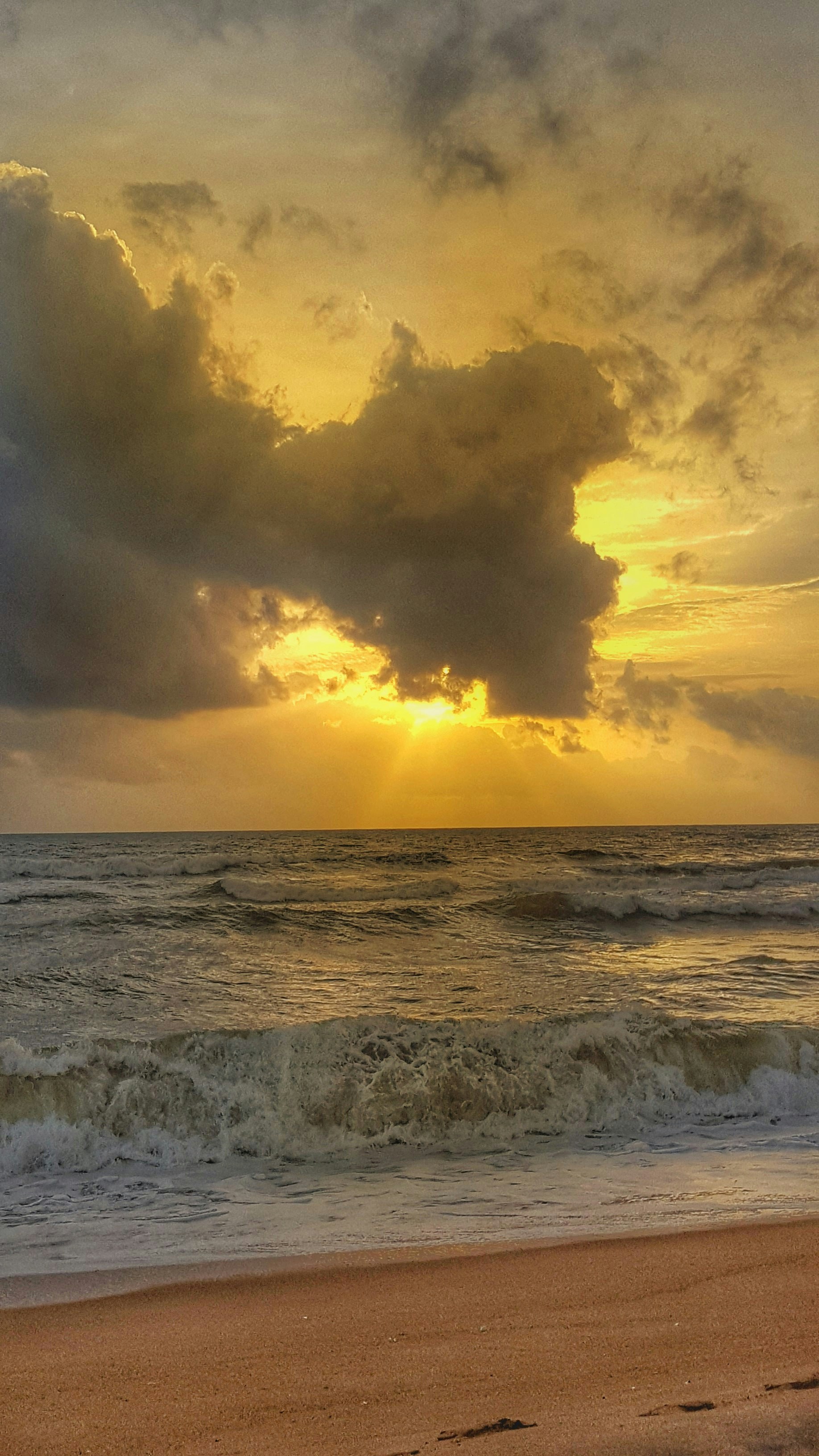 Sunset over the ocean, with dramatic clouds illuminated by golden light and gentle waves lapping at the sandy shore.