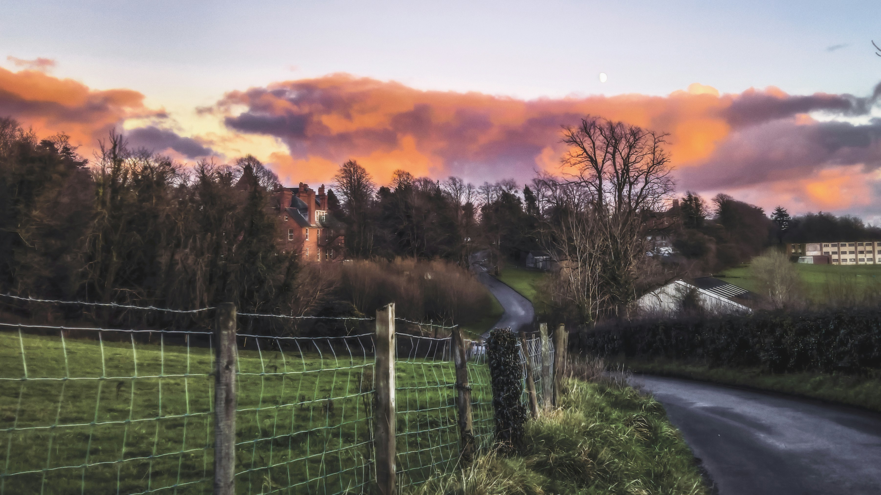 A winding road meanders through lush greenery beneath a sky painted with vibrant twilight hues. The scene captures the serene beauty of rural life.