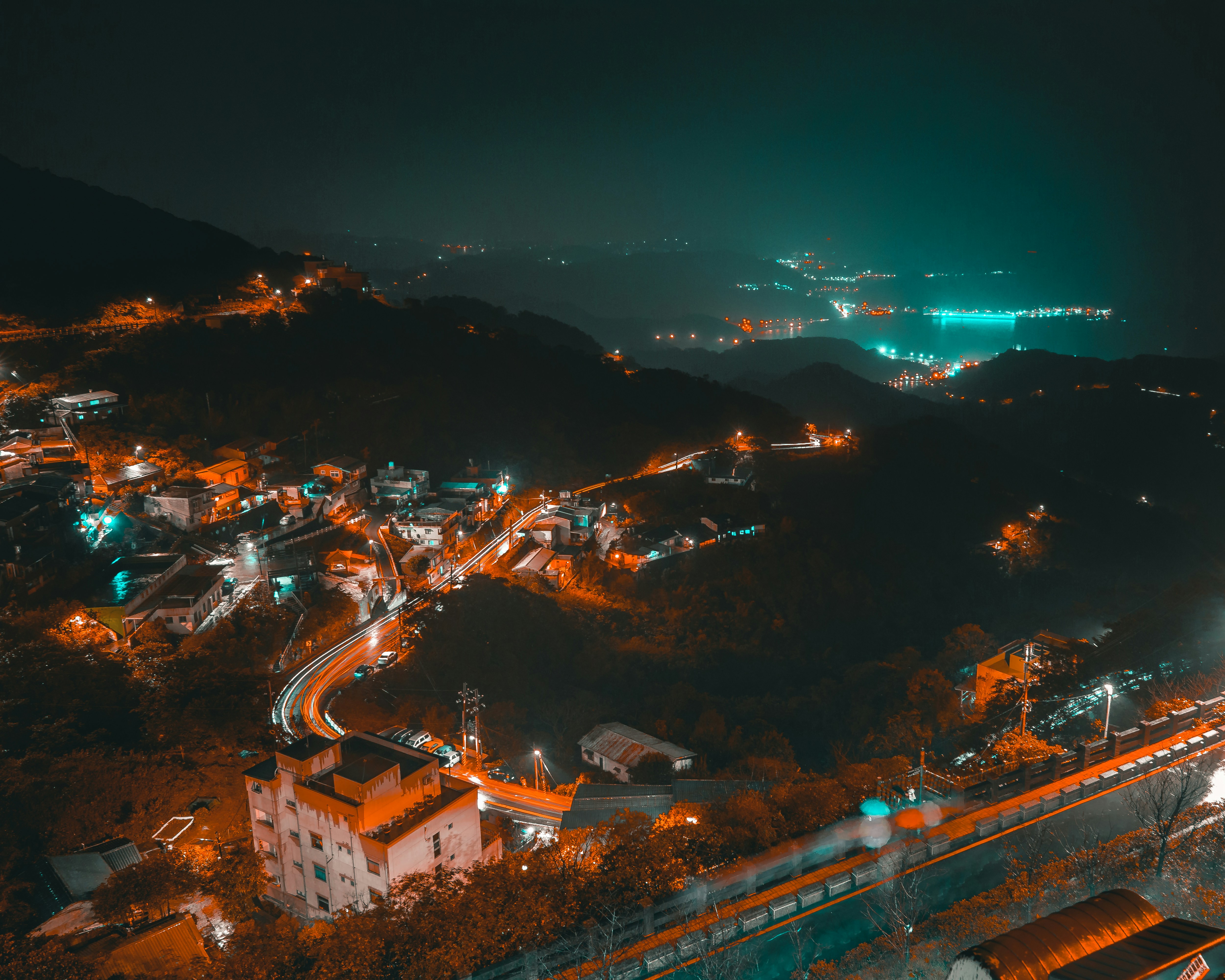Hillside town illuminated by warm streetlights with a distant view of glowing city lights under a night sky.