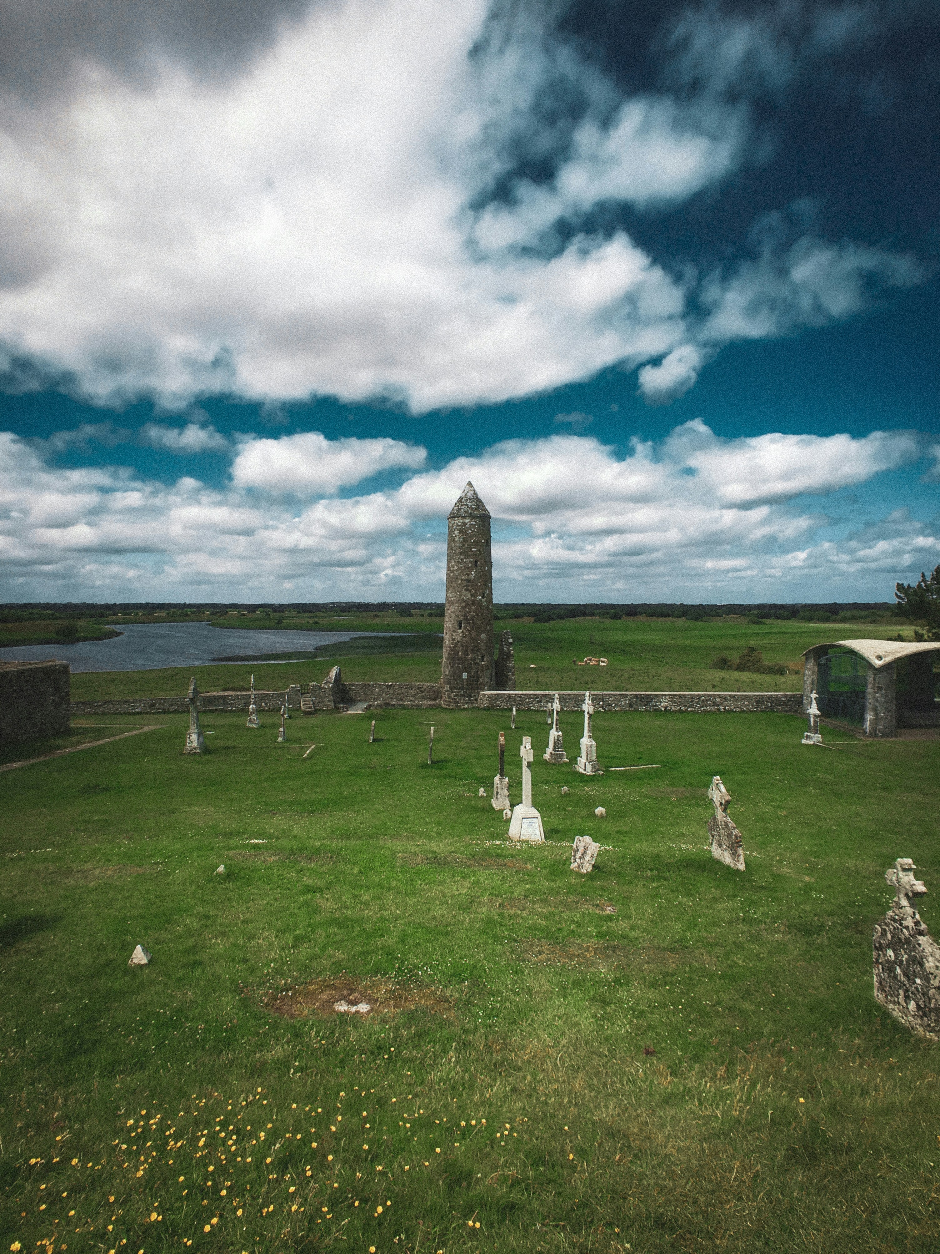 Celtic round tower stands amidst an old graveyard under a vibrant blue sky with scattered clouds.