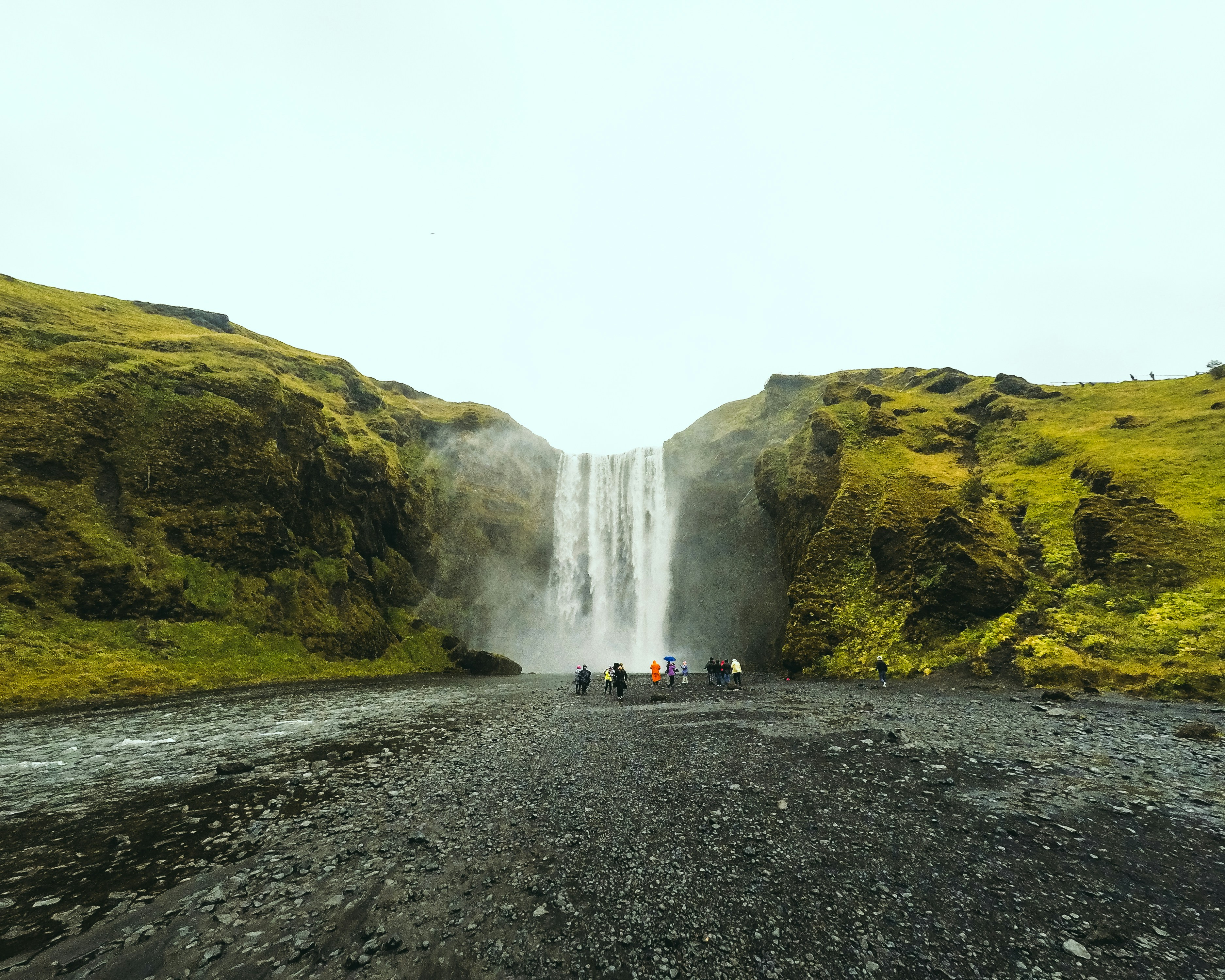 Tourists approach a towering waterfall flanked by lush green cliffs under a cloudy sky.