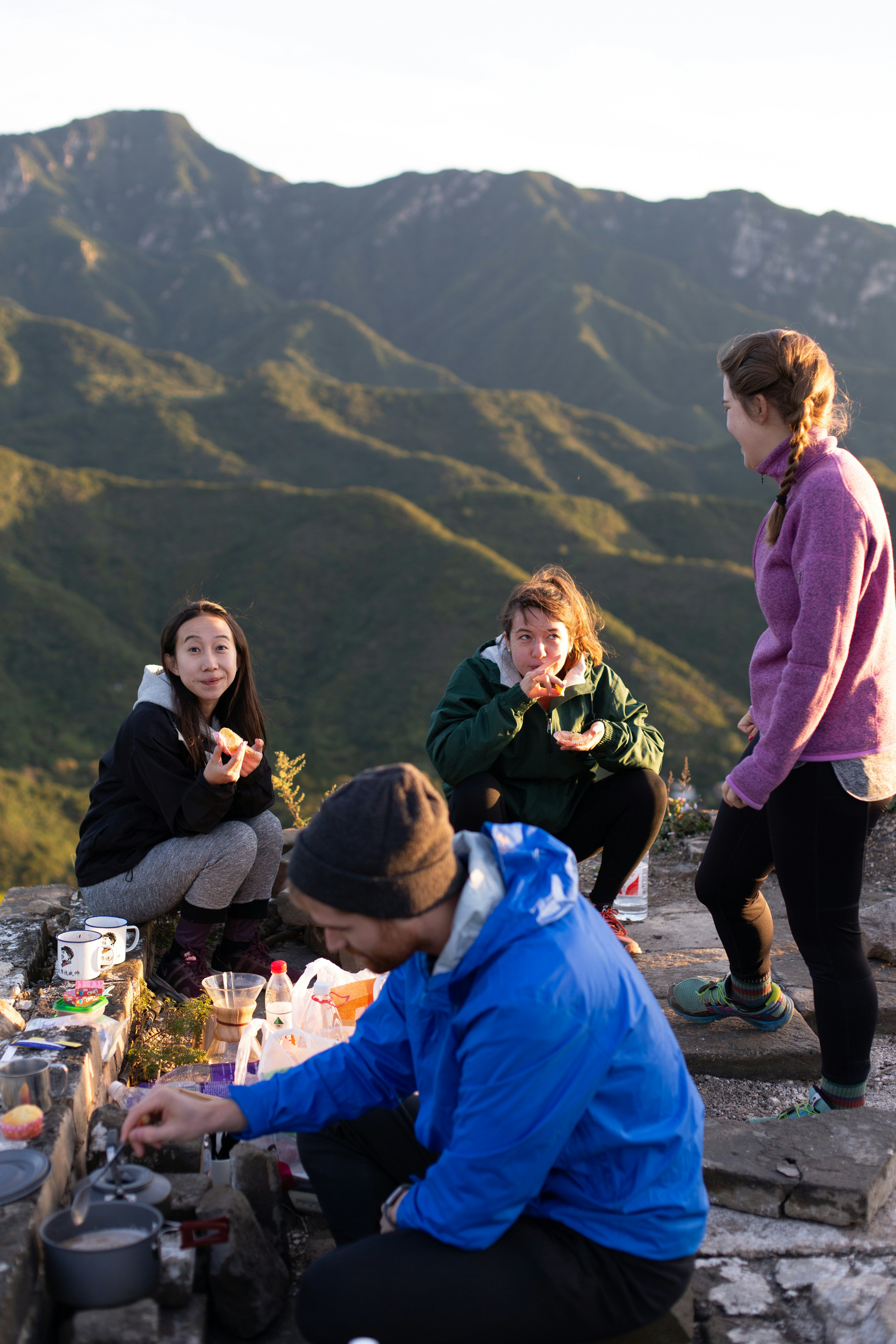 Group of friends enjoying a meal on a mountain ledge, surrounded by lush green hills and distant peaks at sunset.