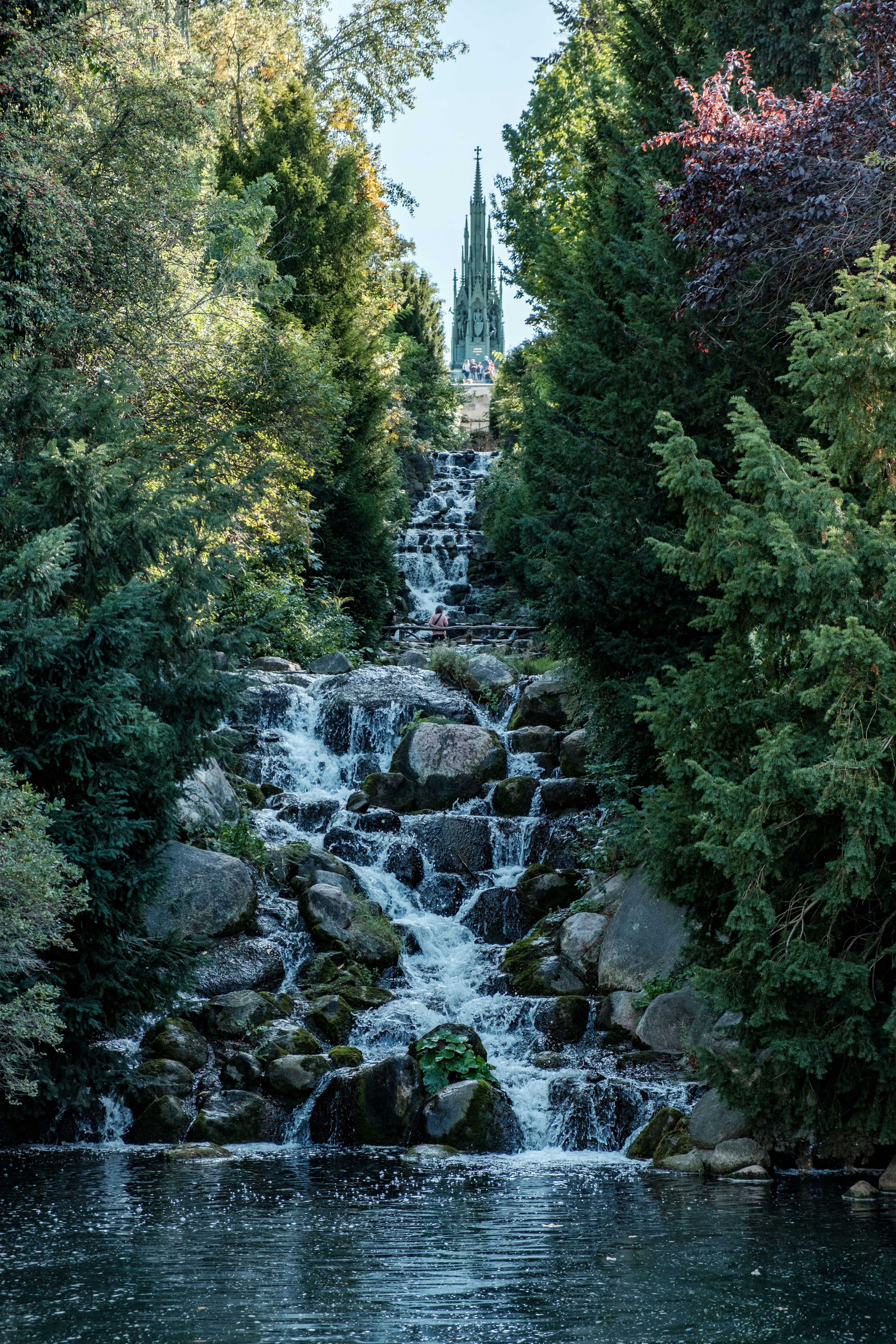 A tranquil waterfall cascades through rocky terrain, framed by lush greenery, with a distant church spire peeking through the trees.