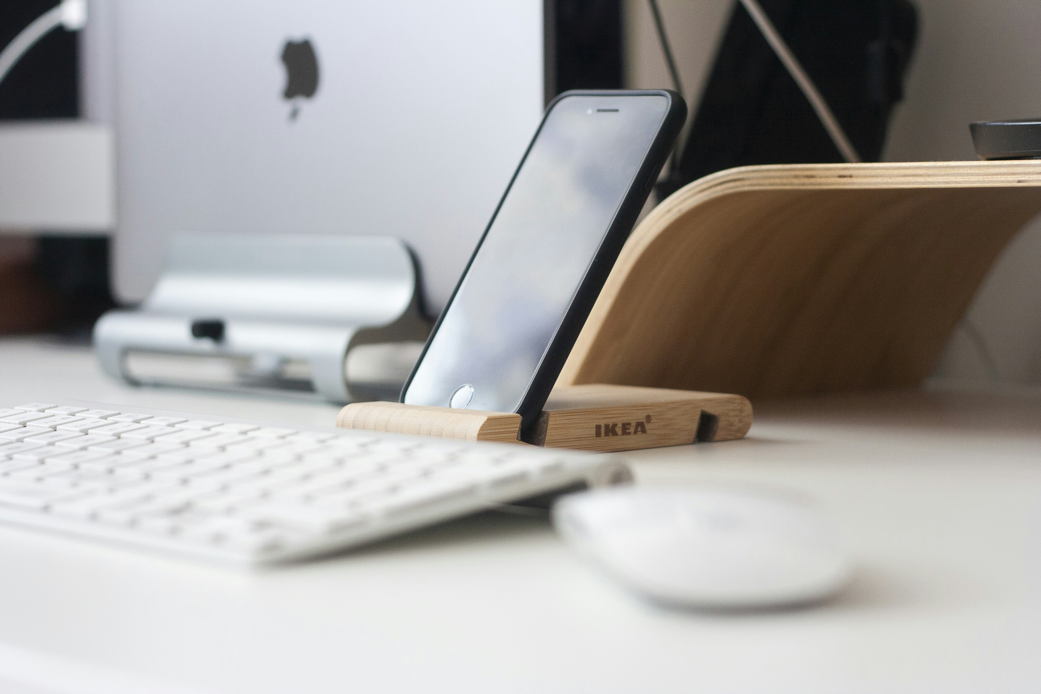 Apple keyboard and mouse on a minimalist desk with a smartphone resting on a wooden stand.