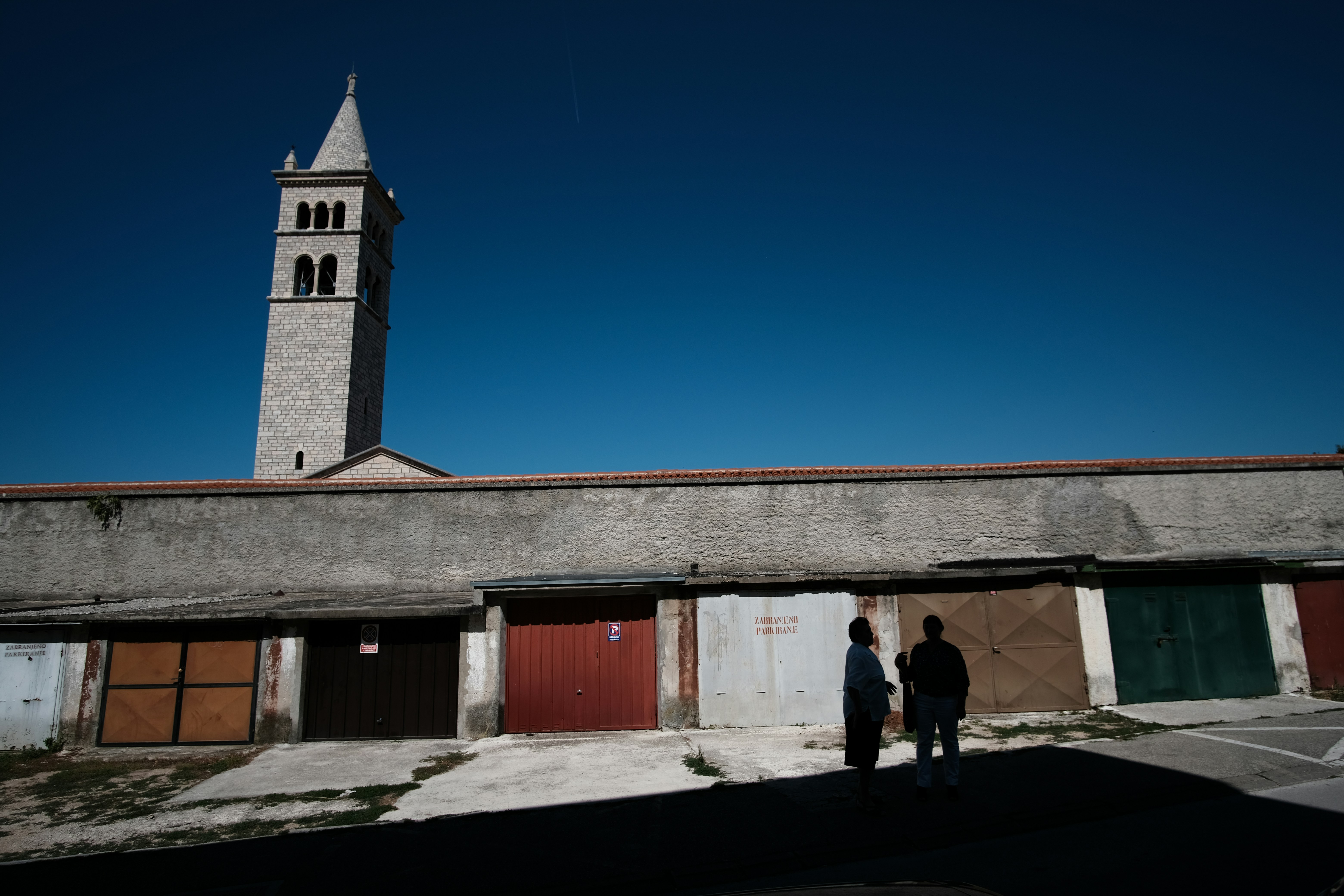 Silhouetted figures converse in front of a weathered building, with a stone tower rising against a clear blue sky.