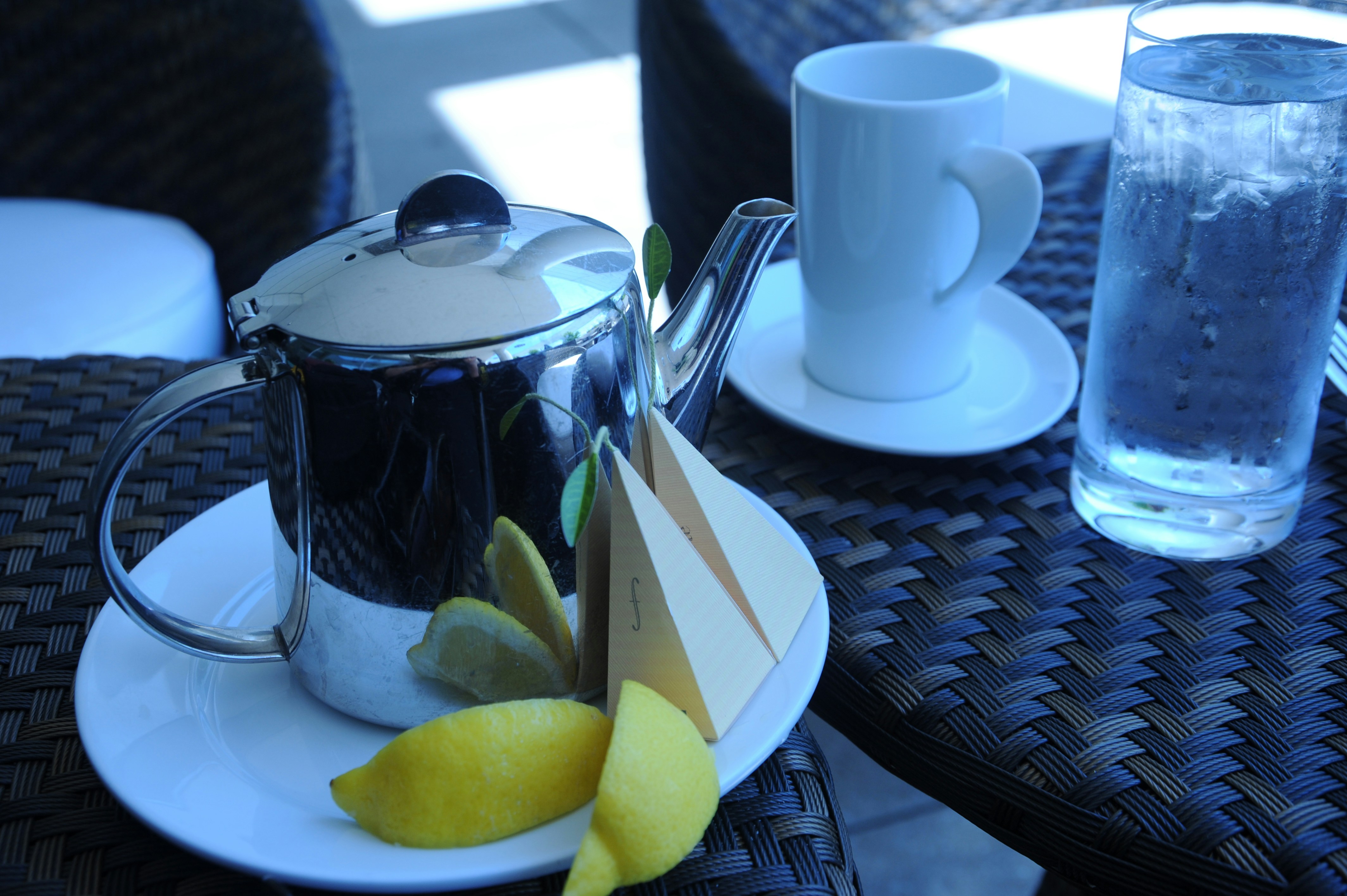 Metallic teapot with lemon slices and triangle tea bags on a white plate, accompanied by a cup and sparkling water on a wicker table.