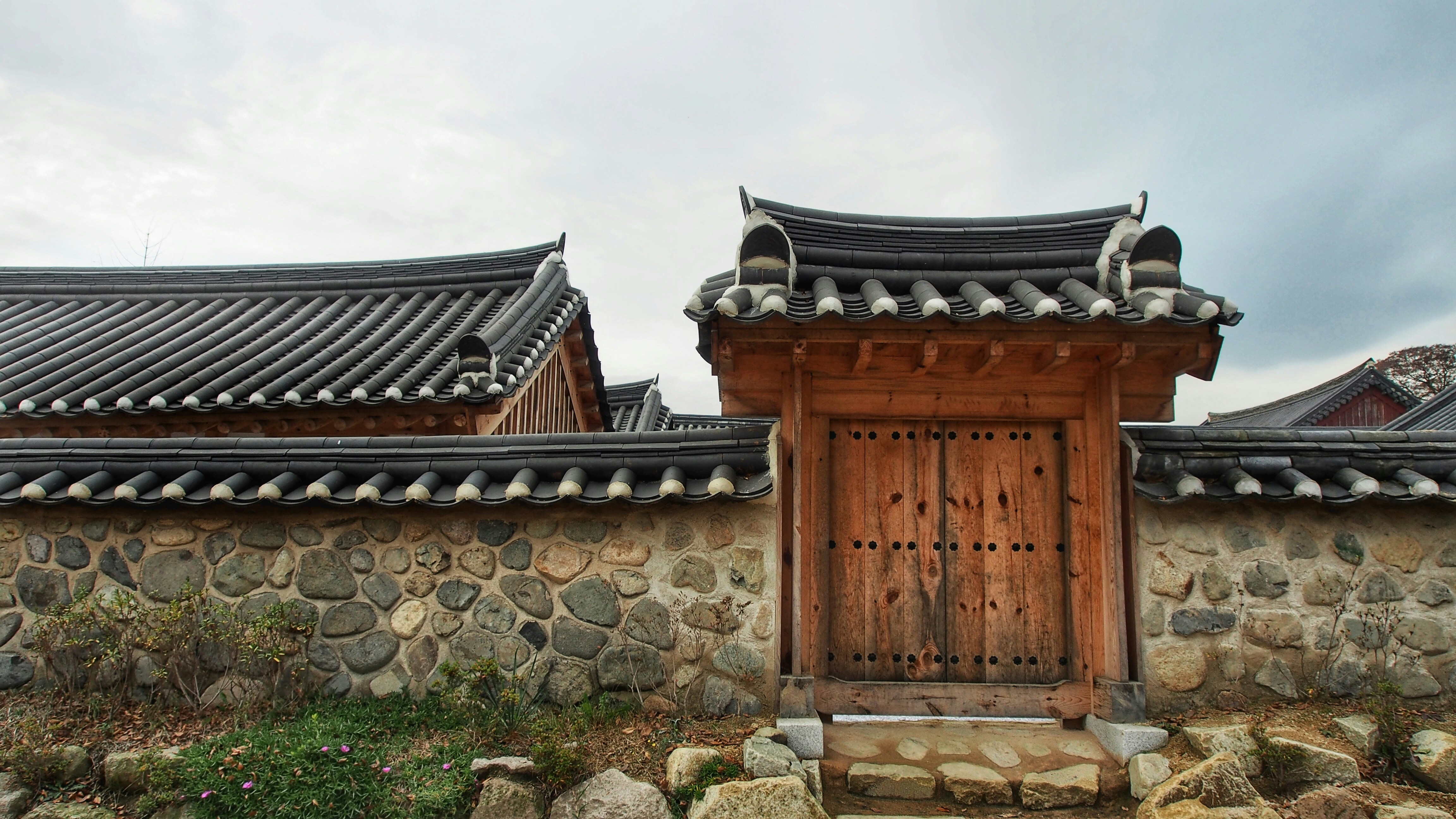 Intricately designed wooden door framed by a stone wall, leading to traditional Korean architecture under a cloudy sky.