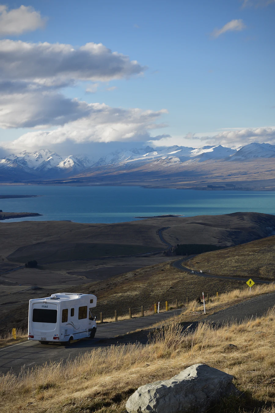 A white camper van descends a winding road through dry hills, with a bright blue lake and snowcapped mountains under scattered clouds in the distance.