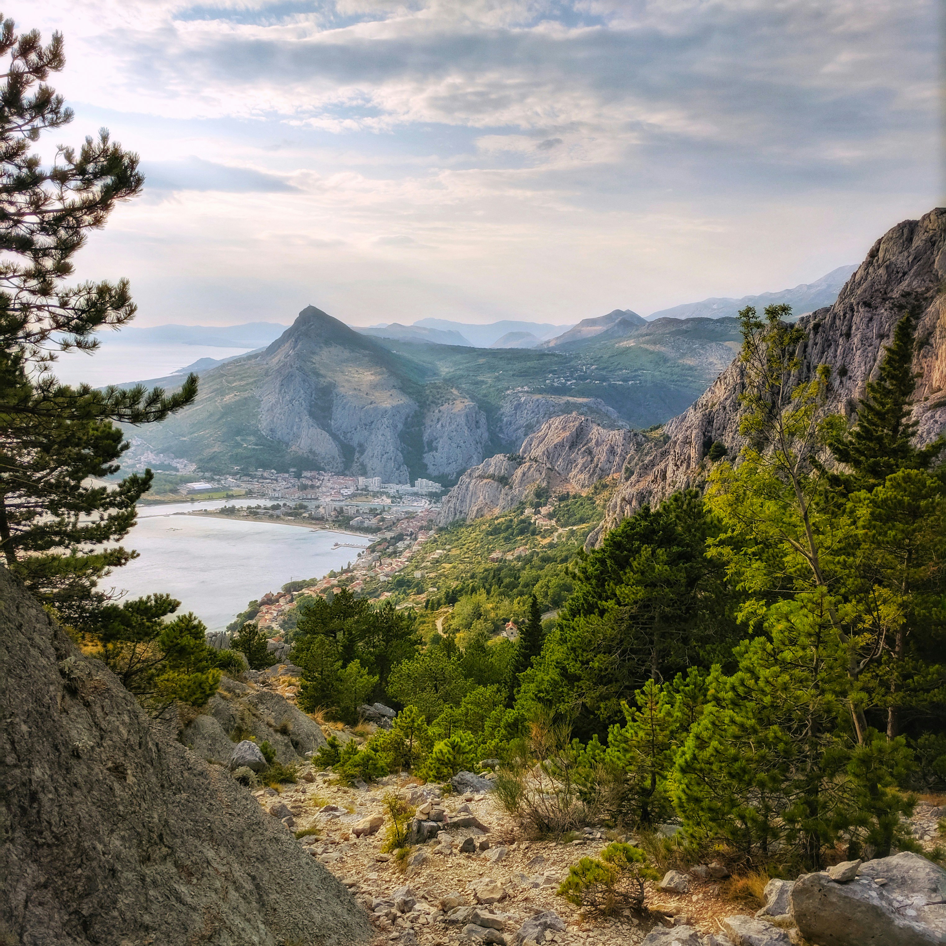 Scenic mountain landscape with rocky cliffs, lush greenery, and a coastal bay under a cloudy sky.