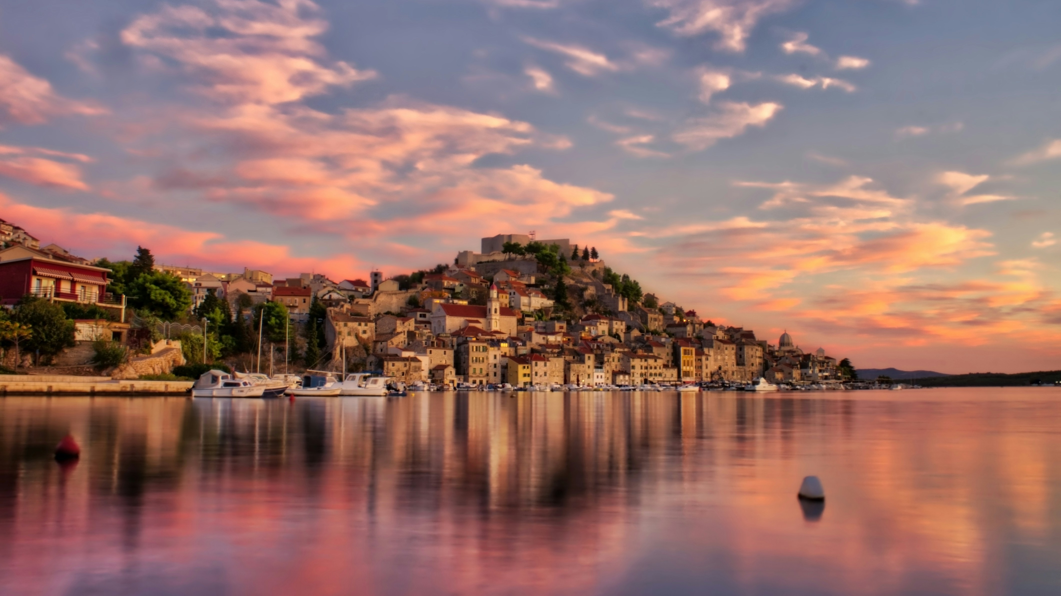 Charming hillside town with colorful buildings overlooking a calm bay at sunset, with boats docked along the waterfront.