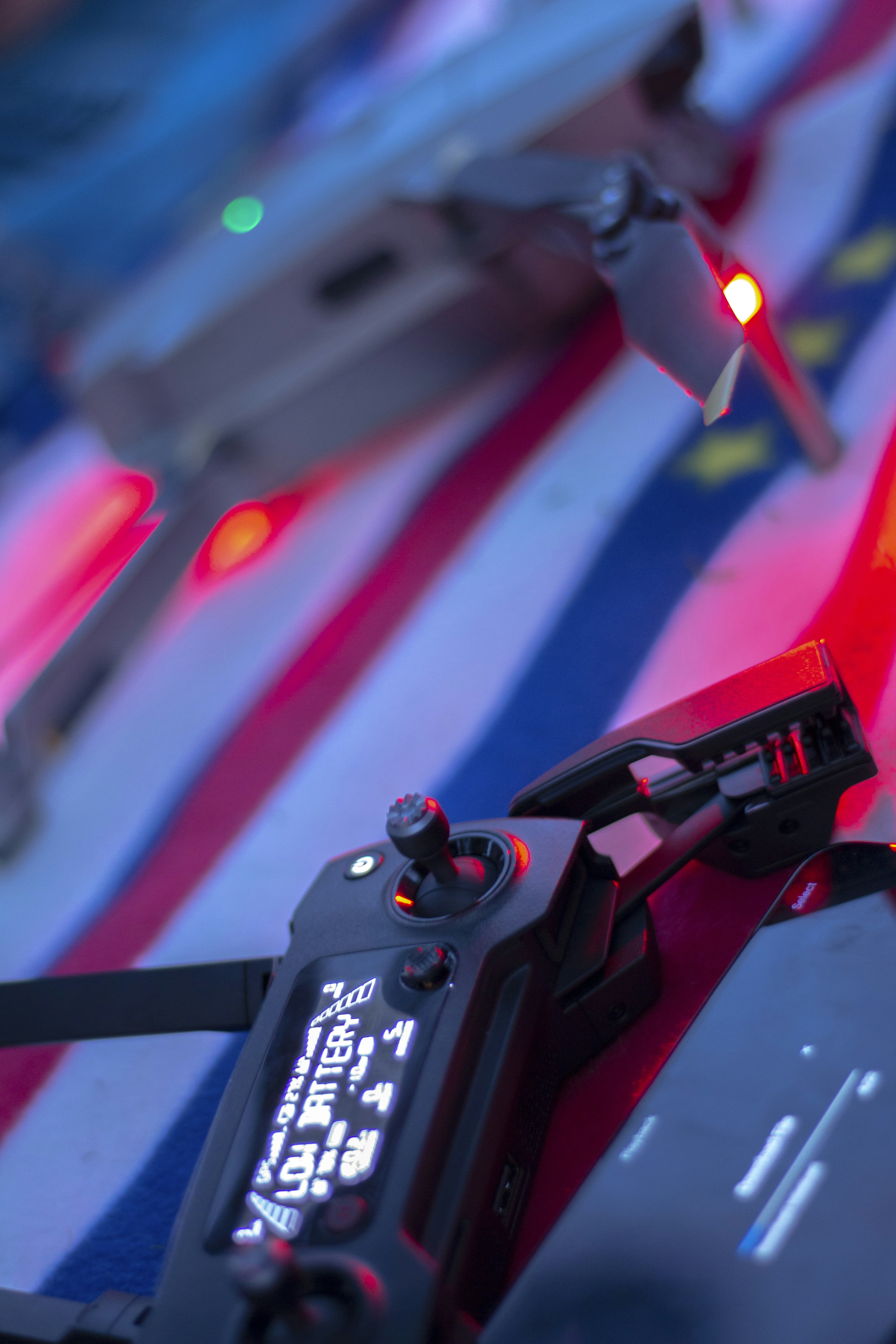 Drone controller with an illuminated display rests on a blue-and-red striped surface bathed in neon lighting.