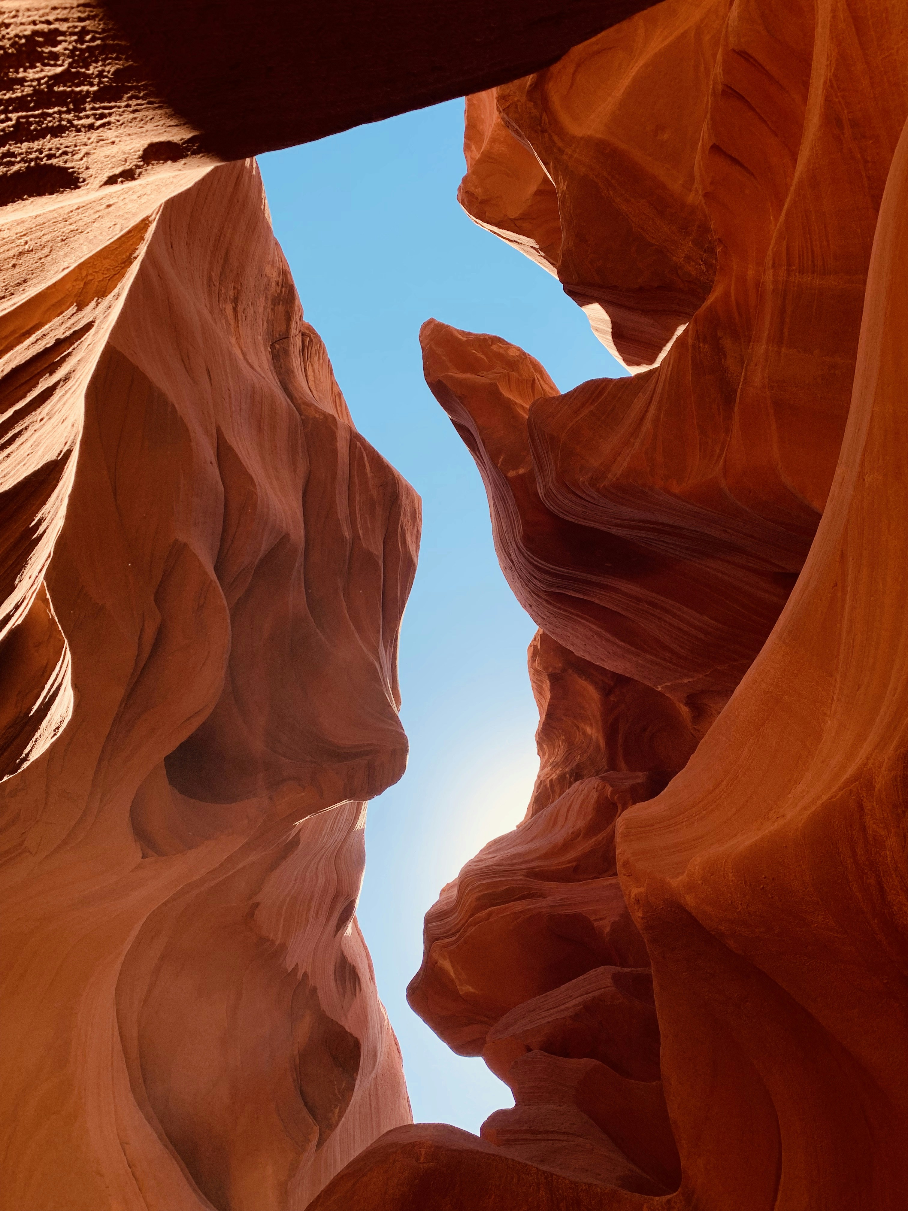 Sunlight filters through the narrow crevices of a sandstone canyon, illuminating the intricate, wave-like formations. The warm hues of the rock create a striking contrast against the blue sky.