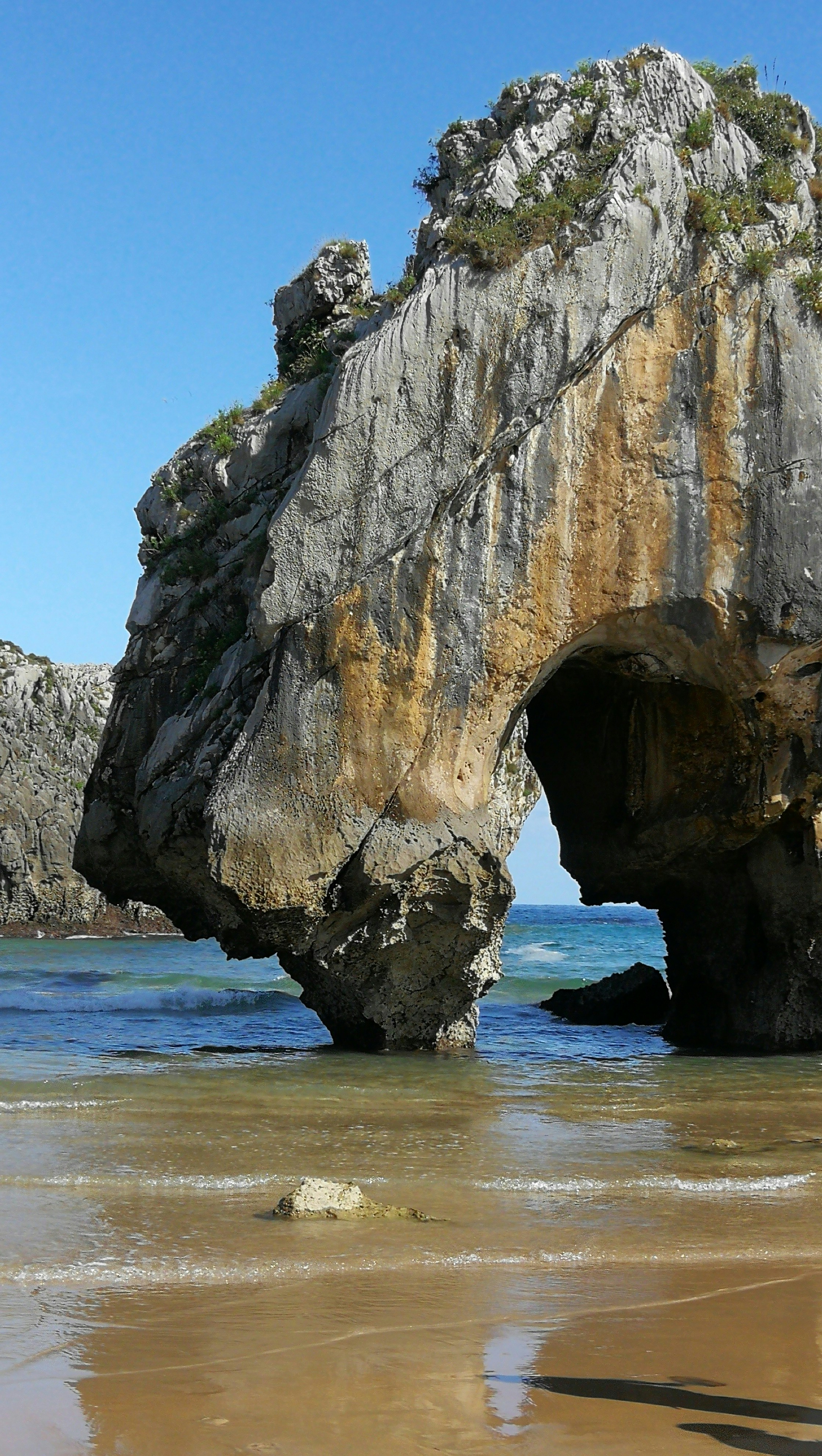A natural rock arch stands majestically on a sandy beach, with waves lapping at its base under a clear blue sky.