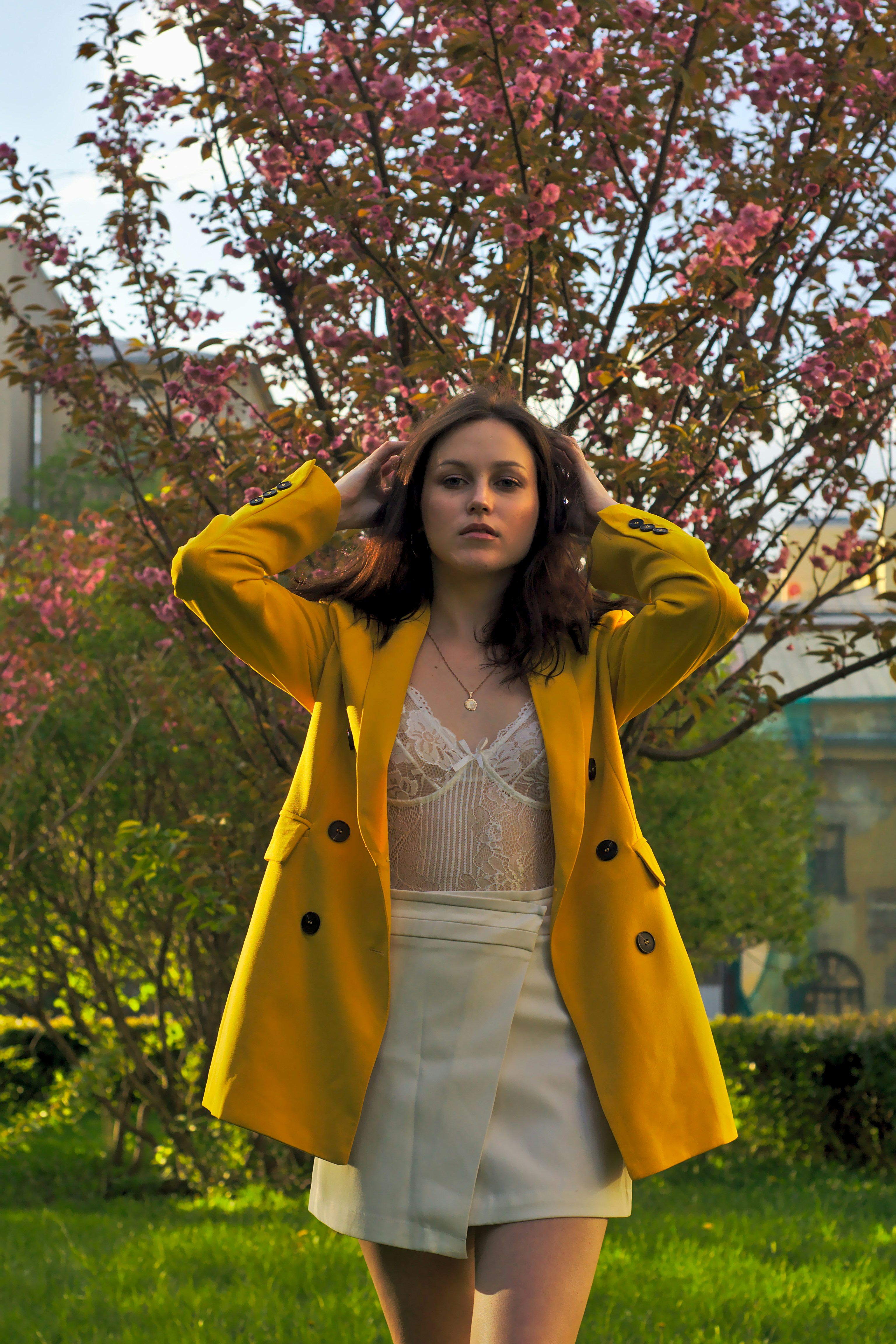 Model in a vibrant yellow coat poses against a backdrop of blooming pink flowers, showcasing a blend of nature and fashion.