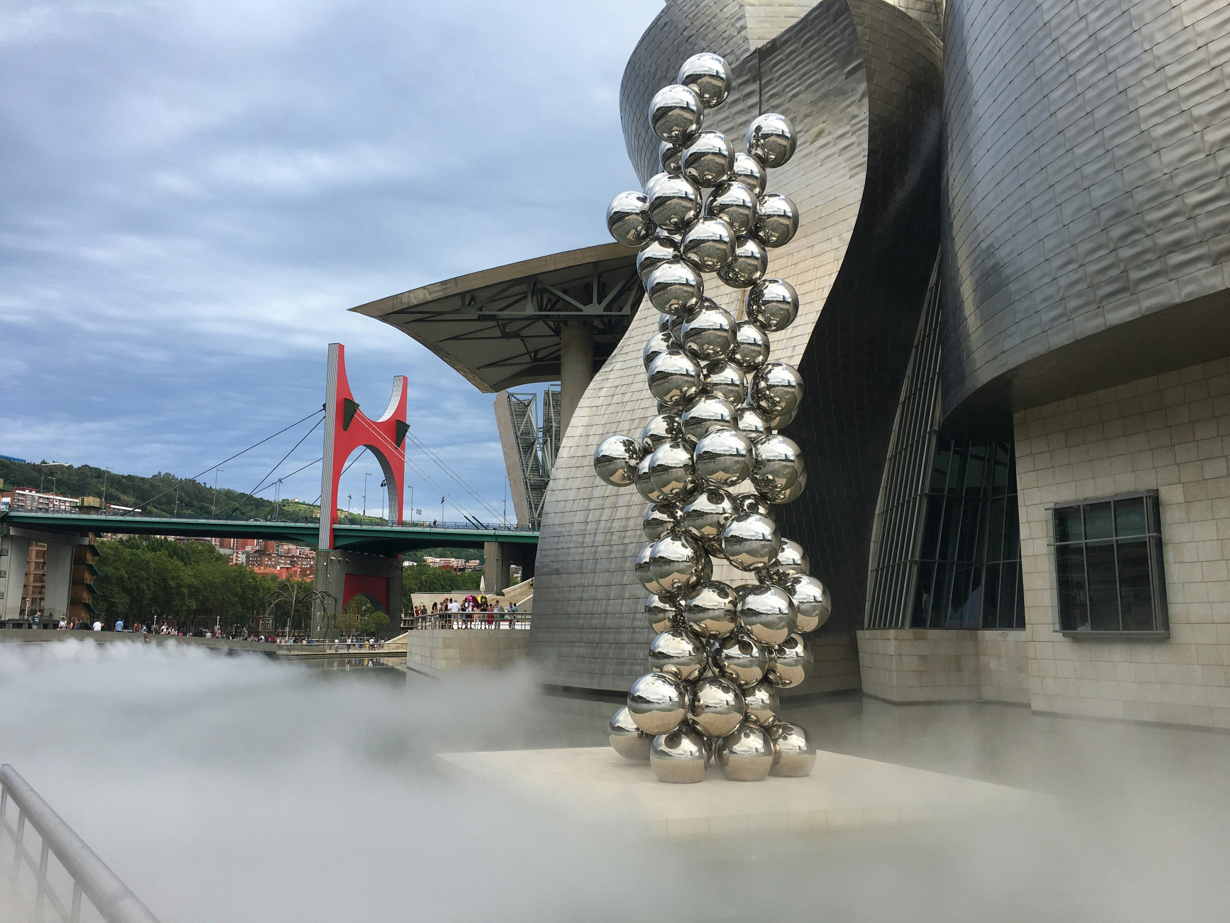 Sculptural installation featuring reflective spheres in front of a contemporary building, with a bridge in the background. Mist surrounds the base, enhancing the visual impact.