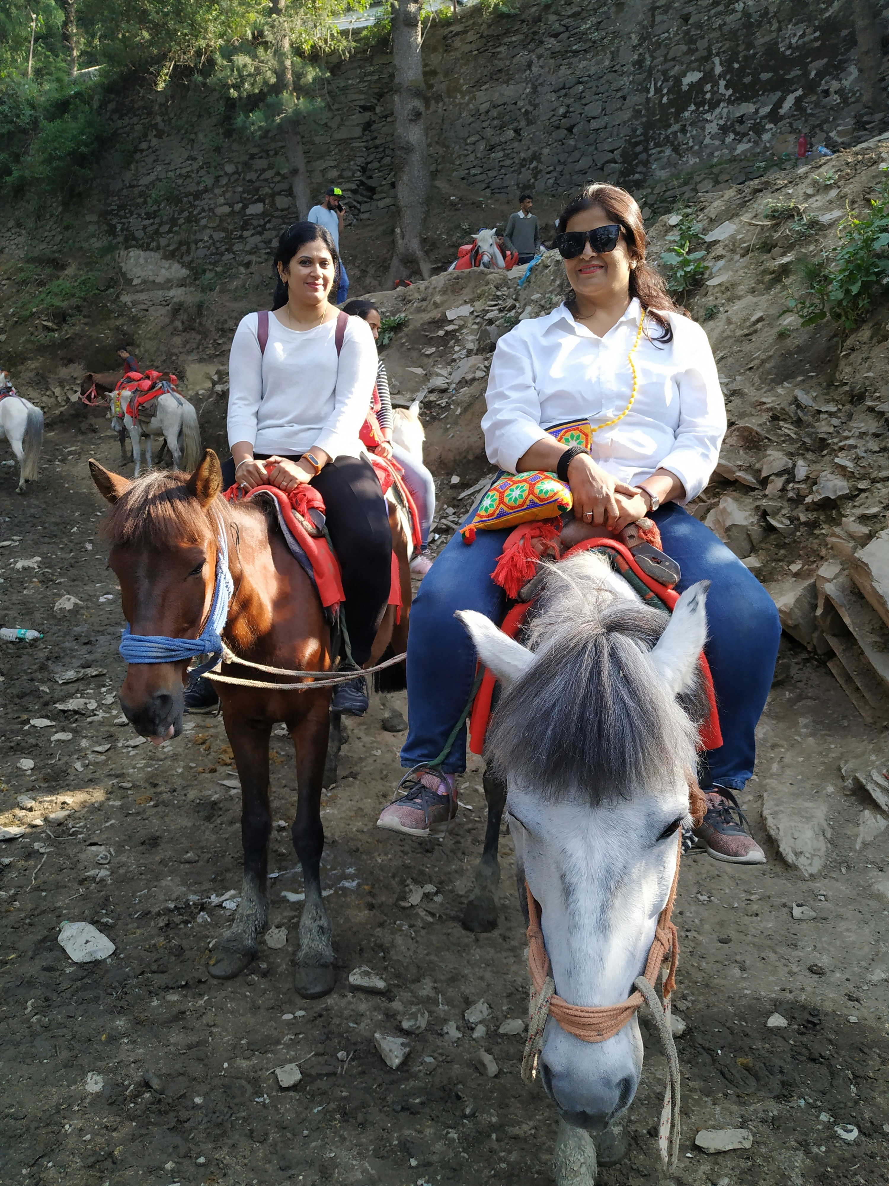 Two women riding horses on a rocky trail, surrounded by greenery and other horses in the background.