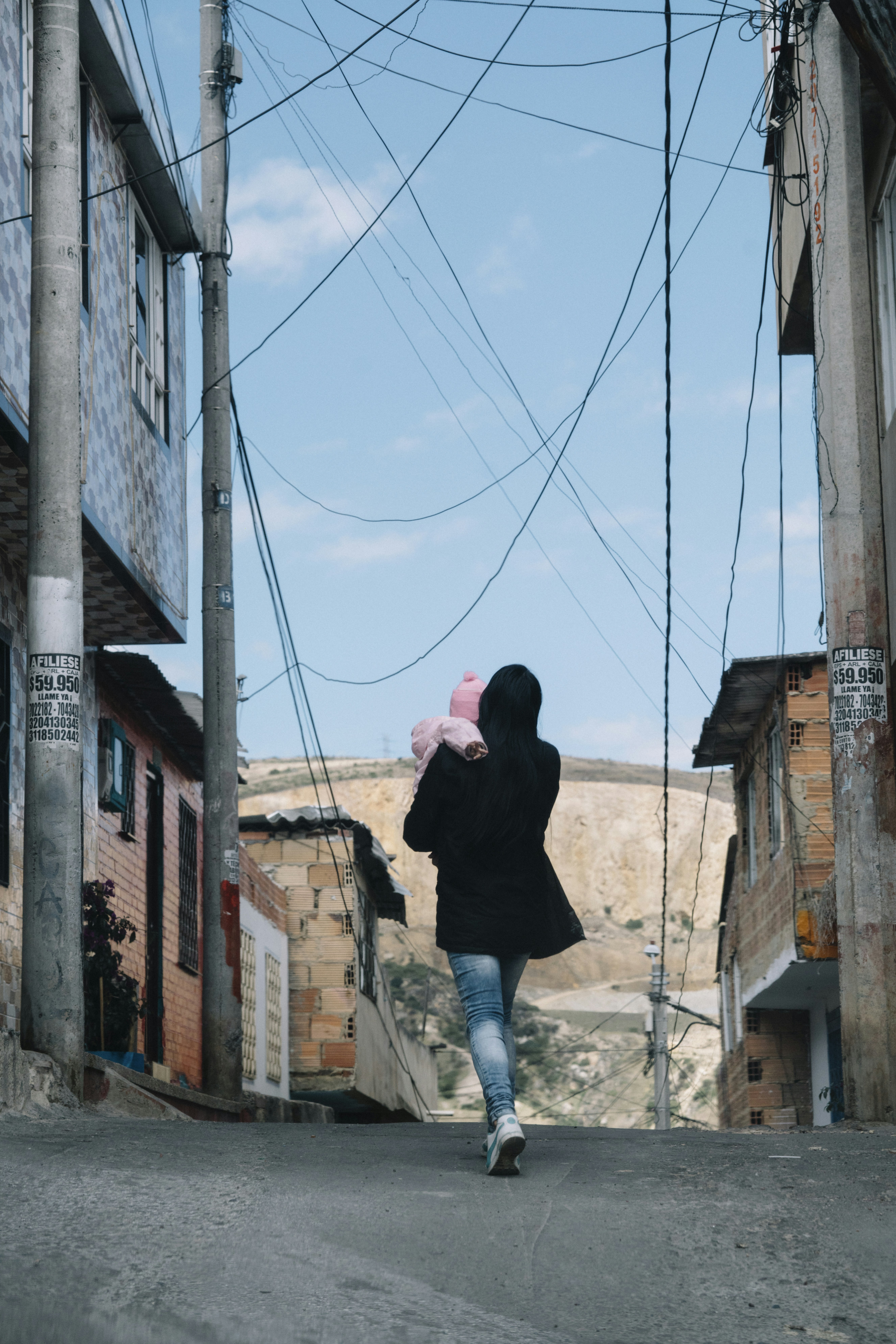 Person walking down a narrow street while carrying a child, surrounded by colorful buildings and power lines. The sky is partially cloudy.