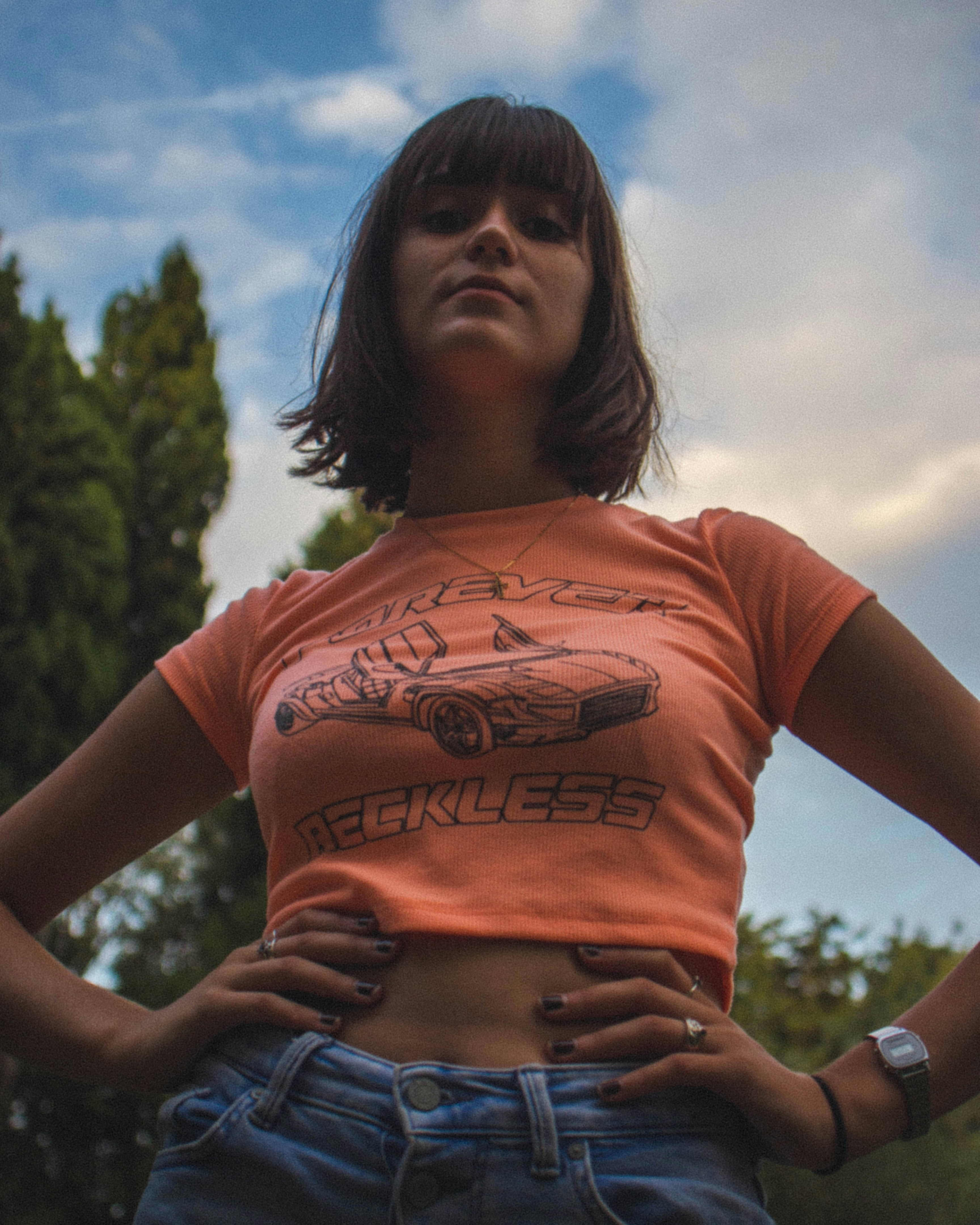 Low-angle portrait of a woman in a coral-orange graphic tee beneath a clear blue sky