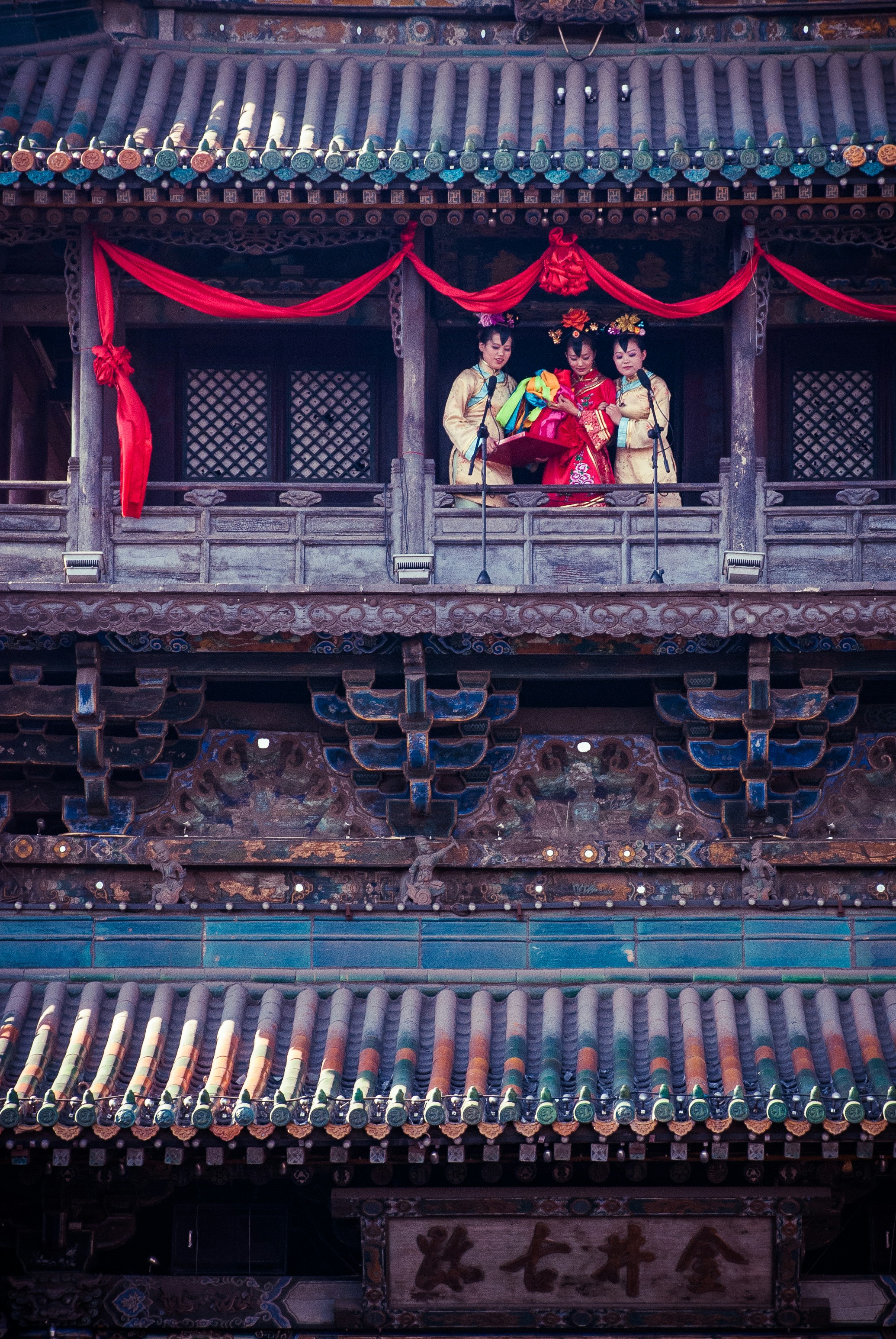 Three individuals dressed in traditional attire stand on a wooden balcony adorned with red ribbons, holding colorful gifts. The intricate architecture showcases cultural artistry.