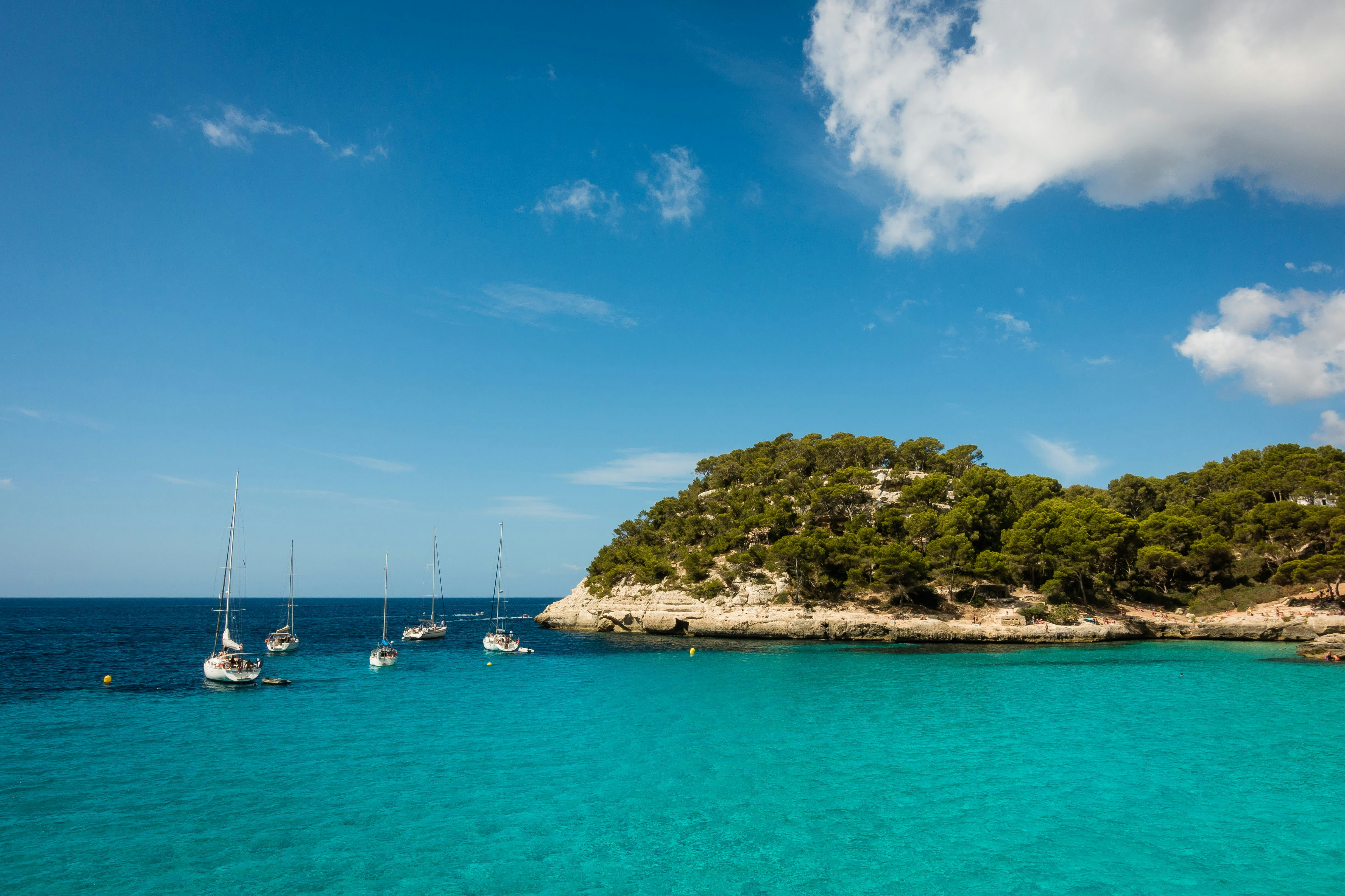 Sailboats anchored in the turquoise waters of Cala Mitjana, framed by lush green cliffs under a bright blue sky.