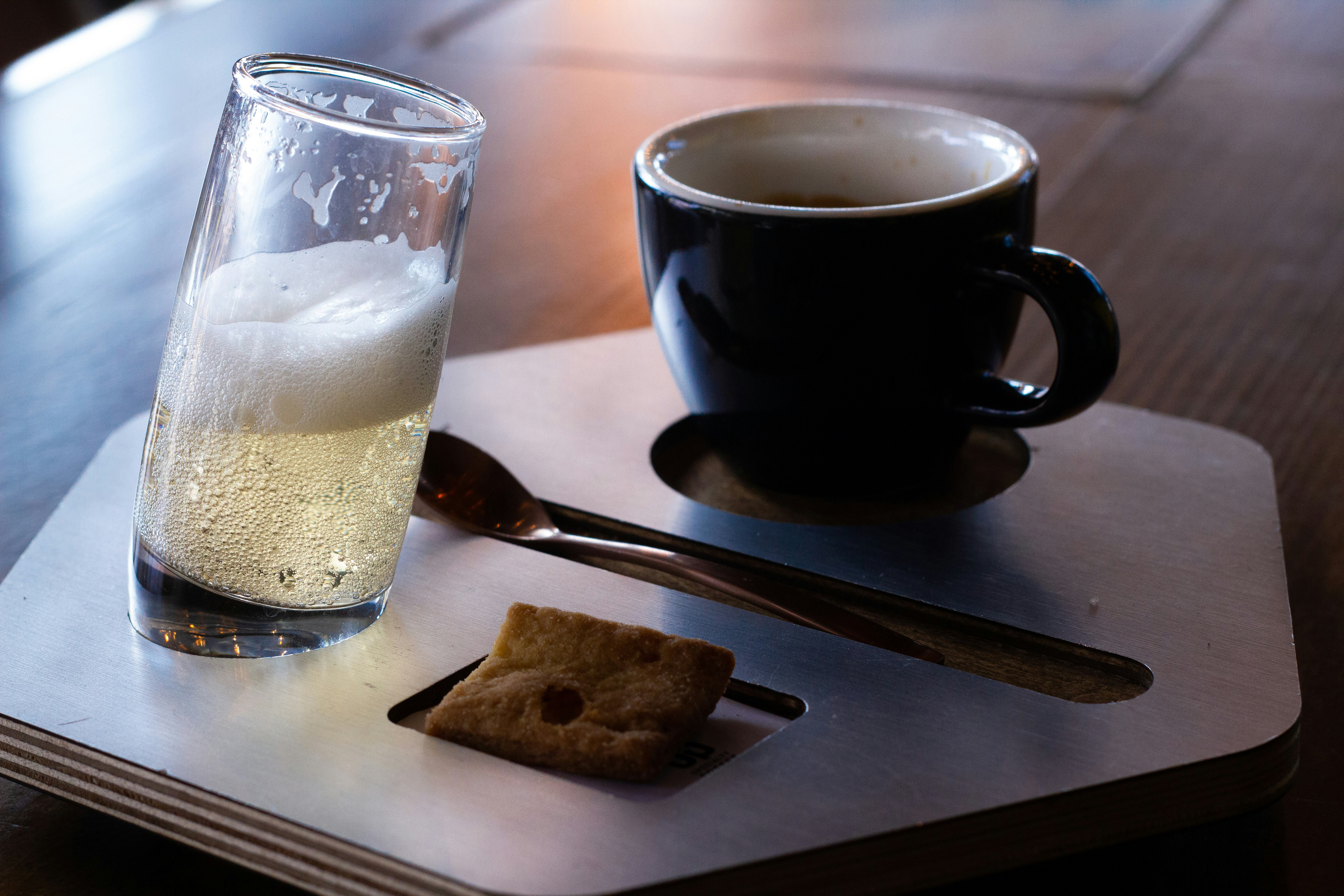 A glass of sparkling beverage stands beside a black coffee cup and a small cookie on a wooden serving tray.