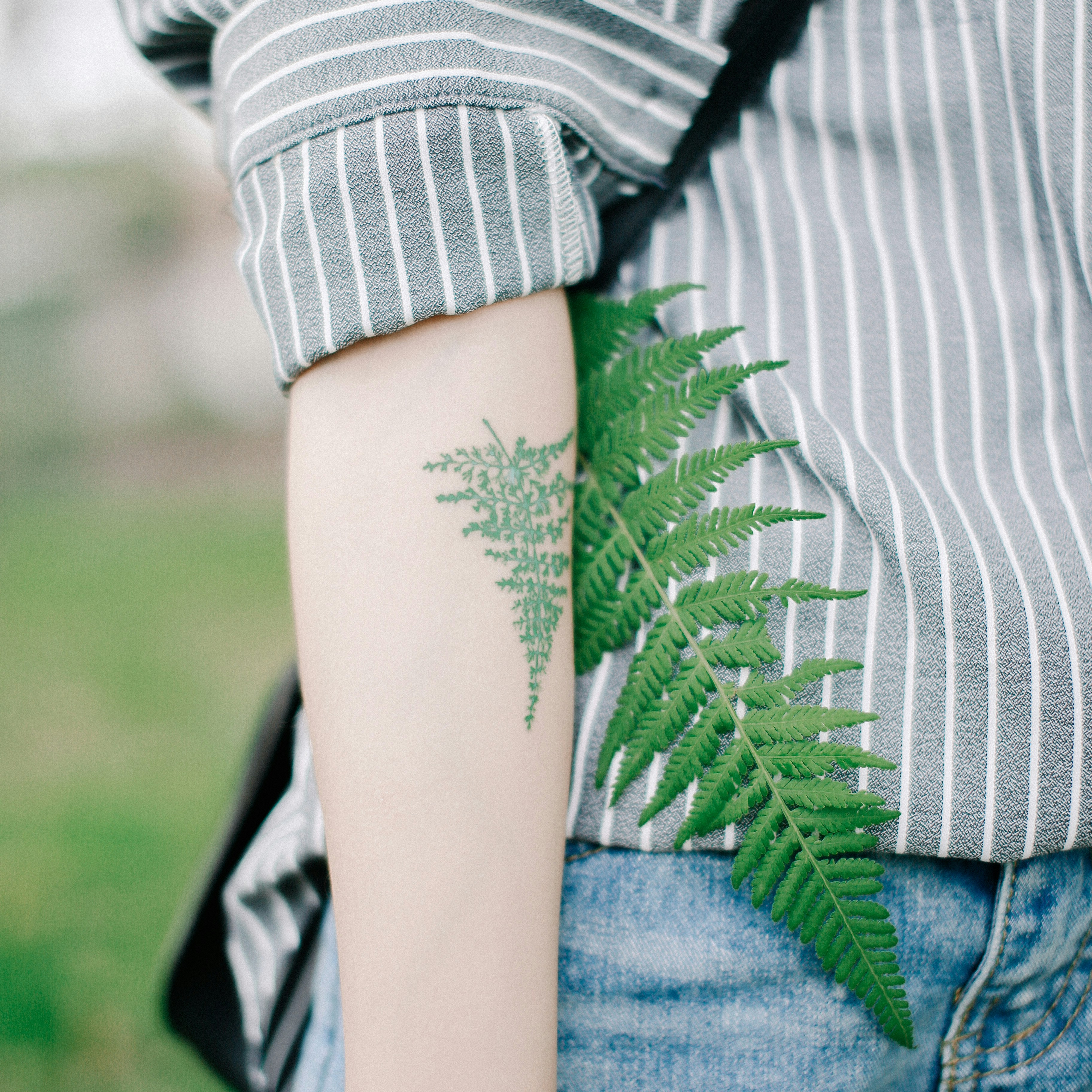 A close-up of a person's arm featuring a green fern tattoo alongside a real fern, set against a blurred natural background.